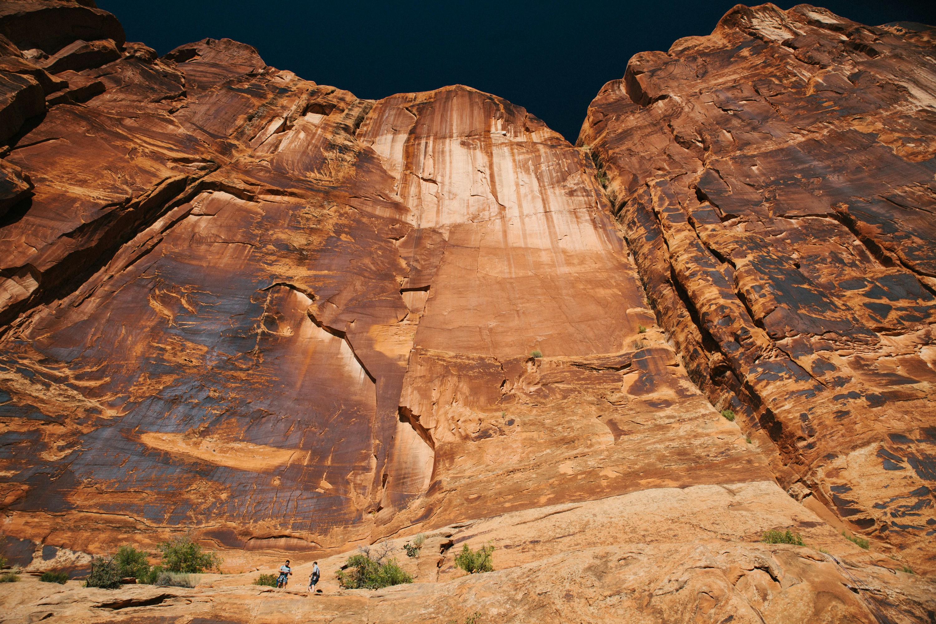 Two climbers navigating the base of towering red rock cliffs under a deep blue sky.