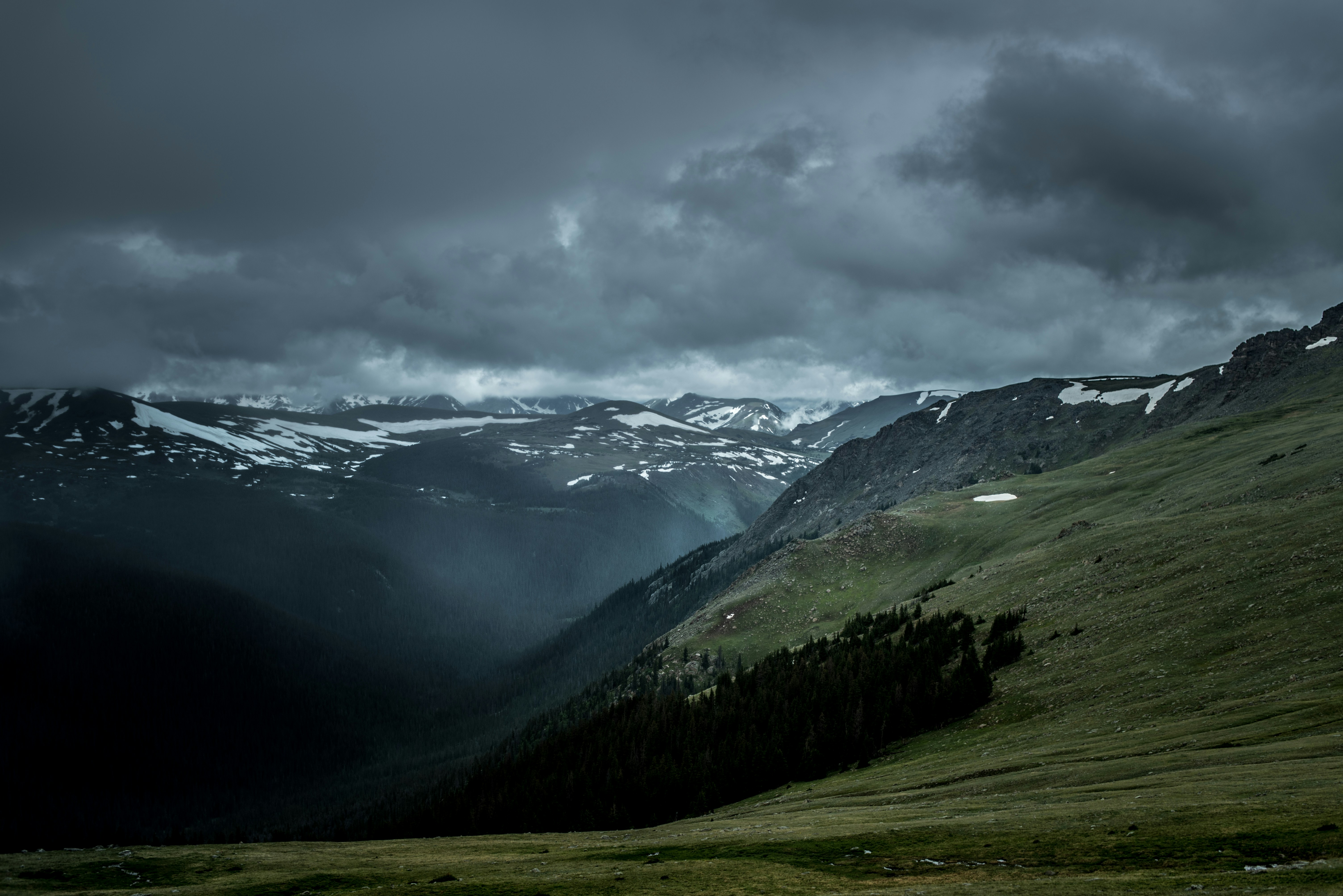 Dramatic landscape showcasing rugged mountains under a moody sky, with patches of snow and lush green valleys. The atmosphere evokes a sense of solitude and the raw beauty of nature.