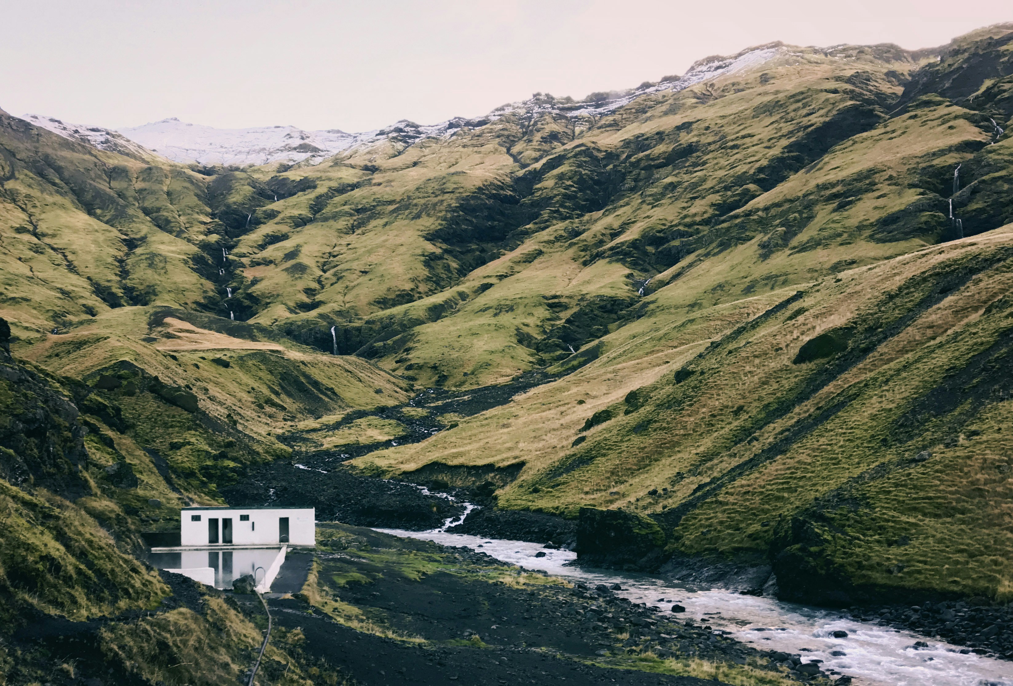 White concrete house by a flowing stream, nestled in lush green mountains.
