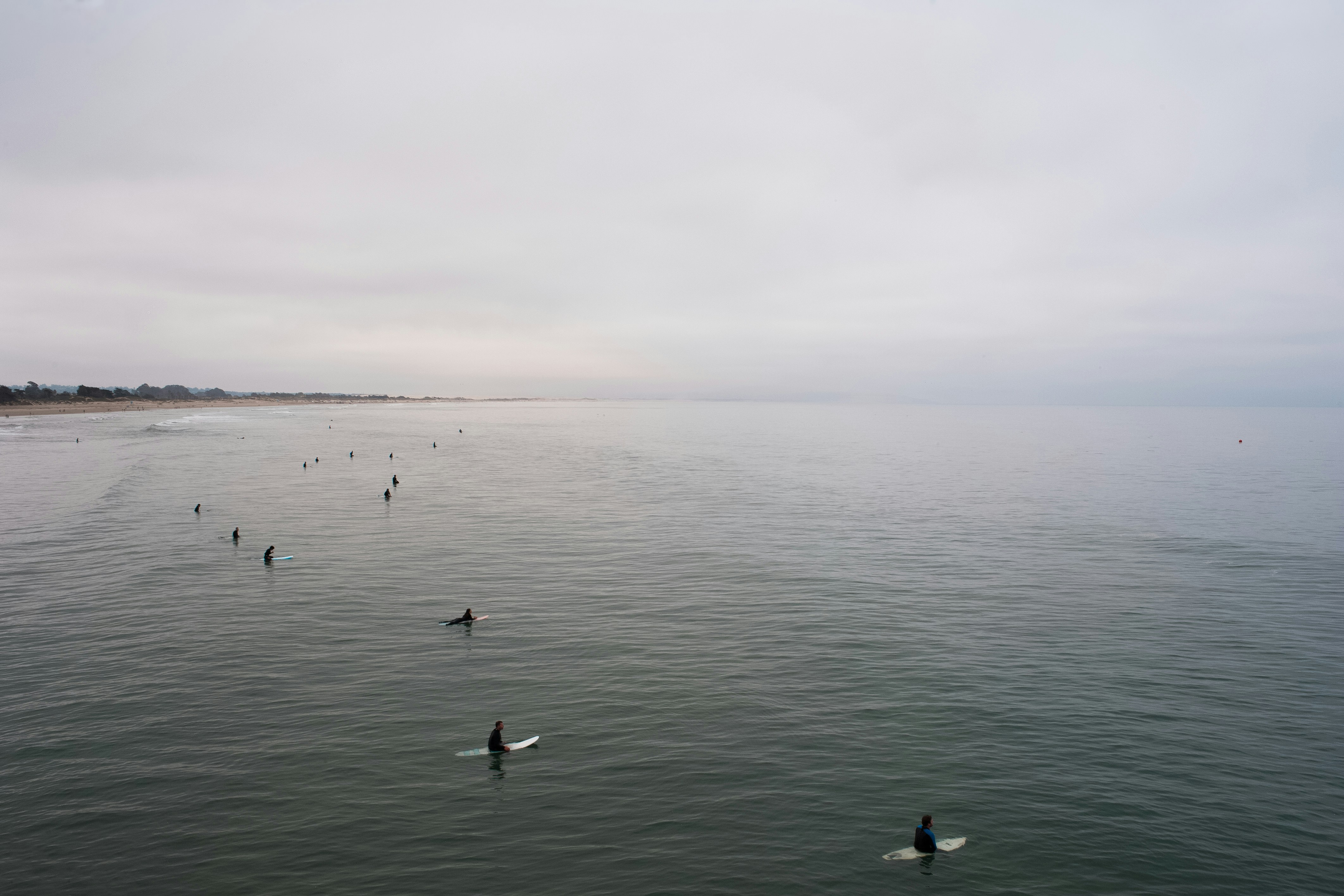 Surfers dot the serene waters, each poised on their boards, as a muted sky looms overhead. The tranquil scene captures the essence of coastal leisure.