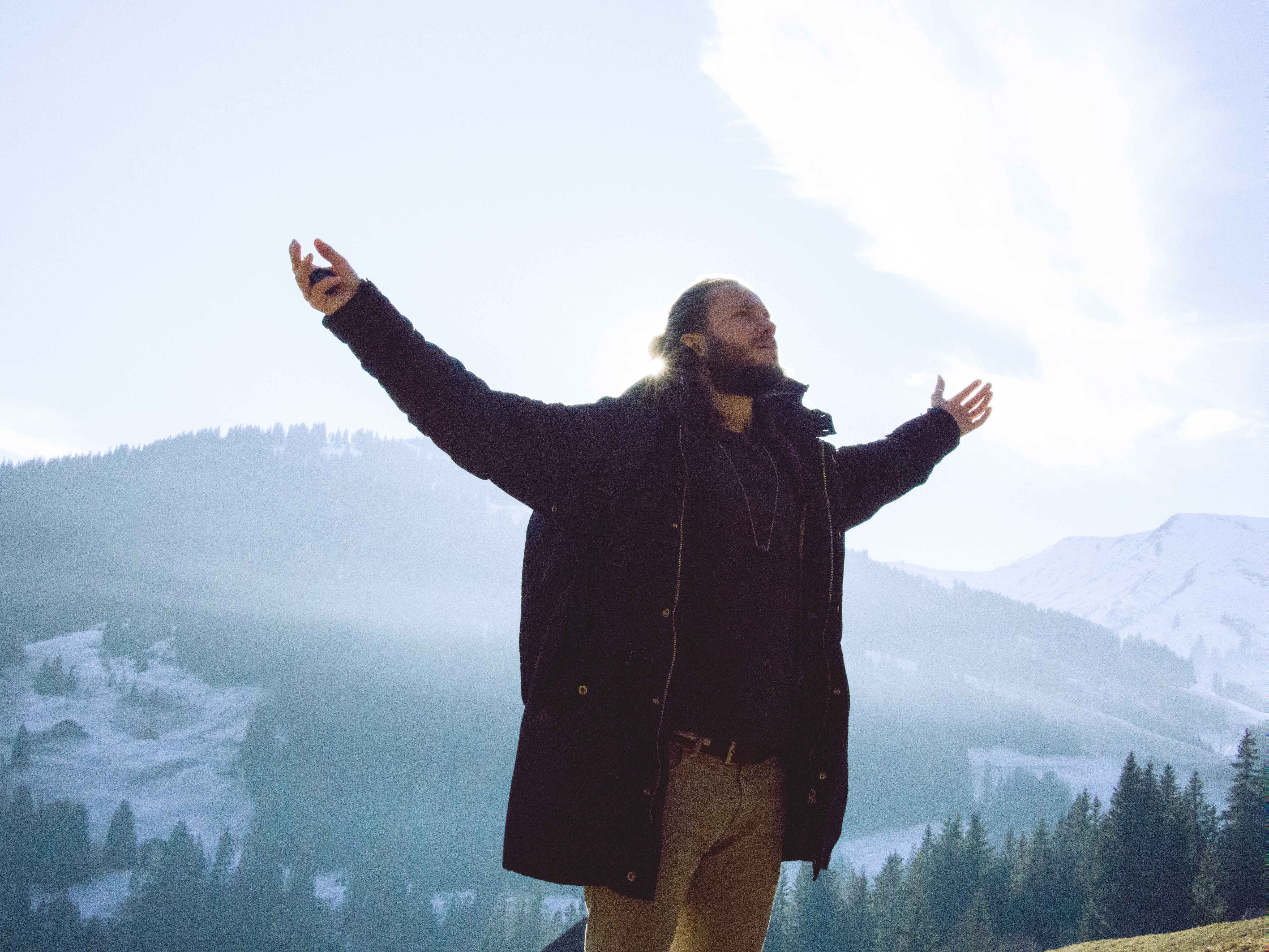 Individual standing with arms outstretched against a backdrop of snow-capped mountains and a clear blue sky.