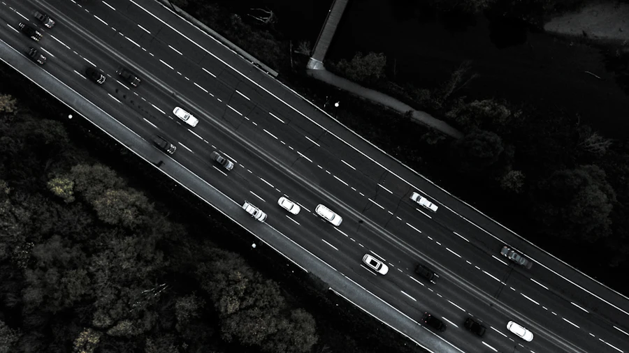 Diagonal highway cutting through dark trees, with a line of cars moving along the lanes at night. High-contrast monochrome look emphasizes the road geometry and traffic flow.