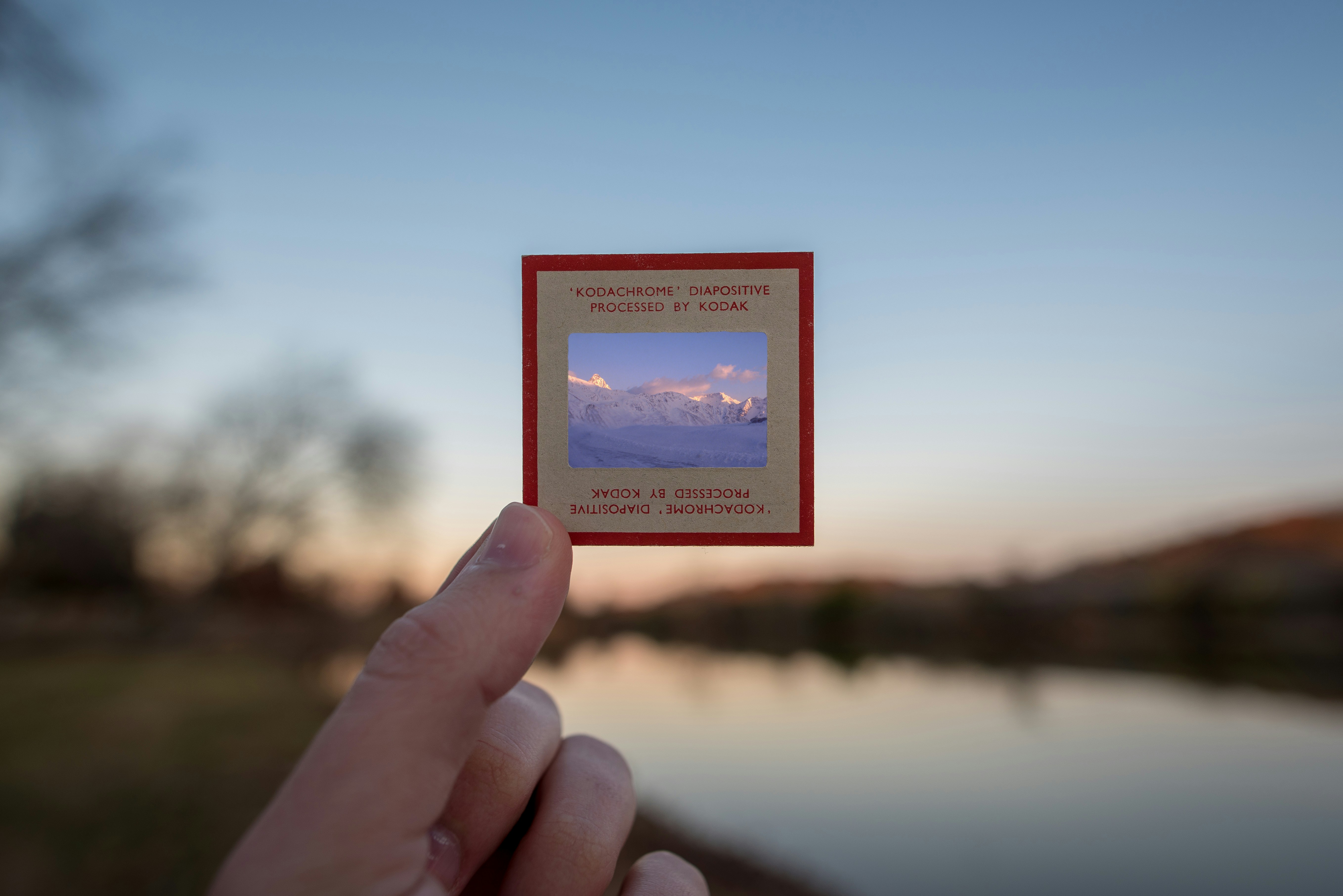 A hand holds a vintage slide against a serene landscape, showcasing majestic mountains and a tranquil river. The scene captures the essence of nostalgia and exploration.