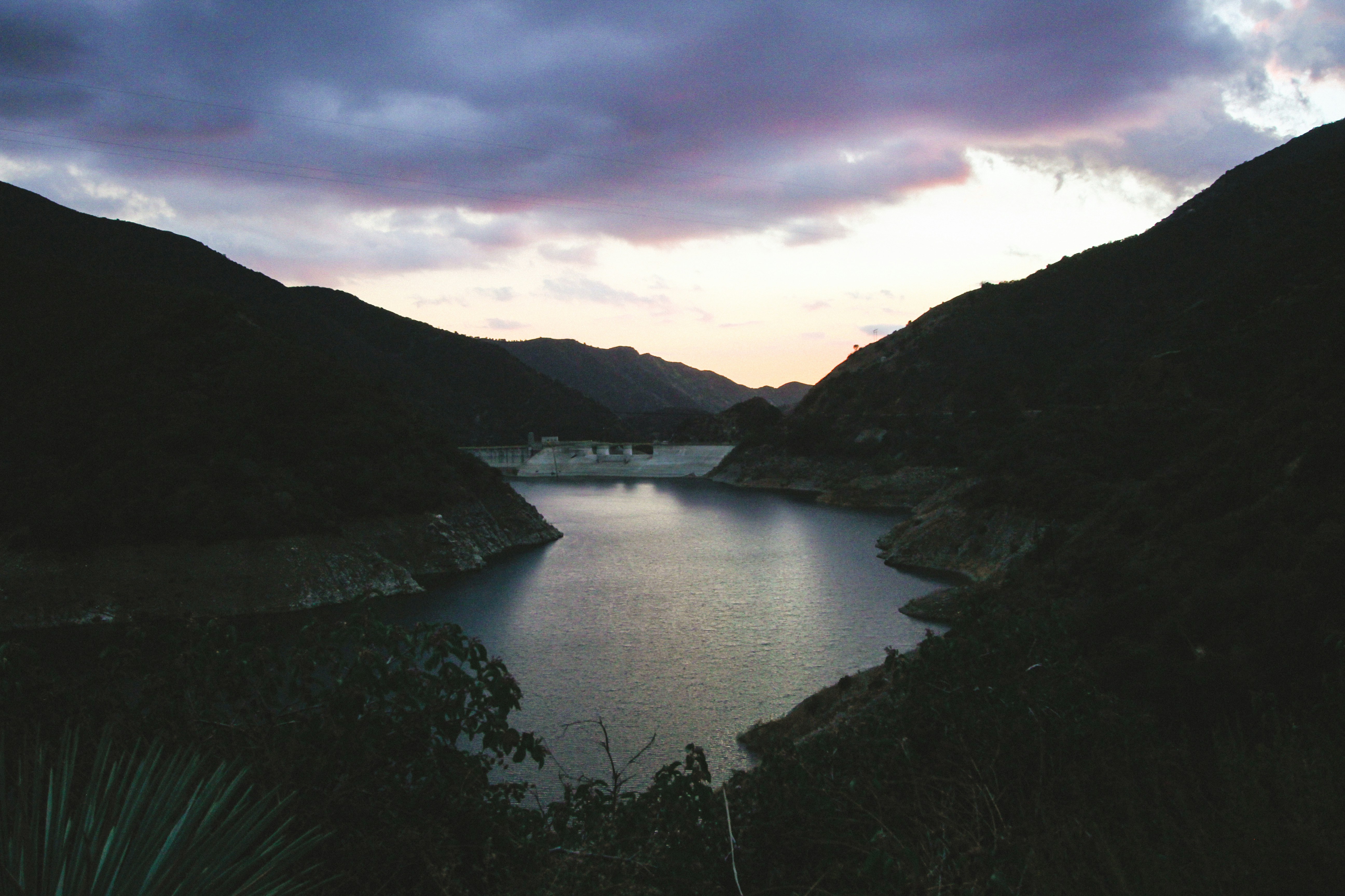 Mountain reservoir under a dramatic sunset sky, flanked by silhouettes of rugged hills.