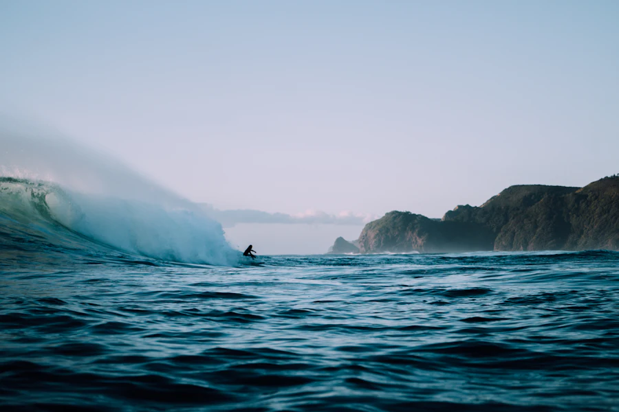A lone surfer rides low inside a breaking blue wave with rocky coastline in the distance and a soft pastel sky above.