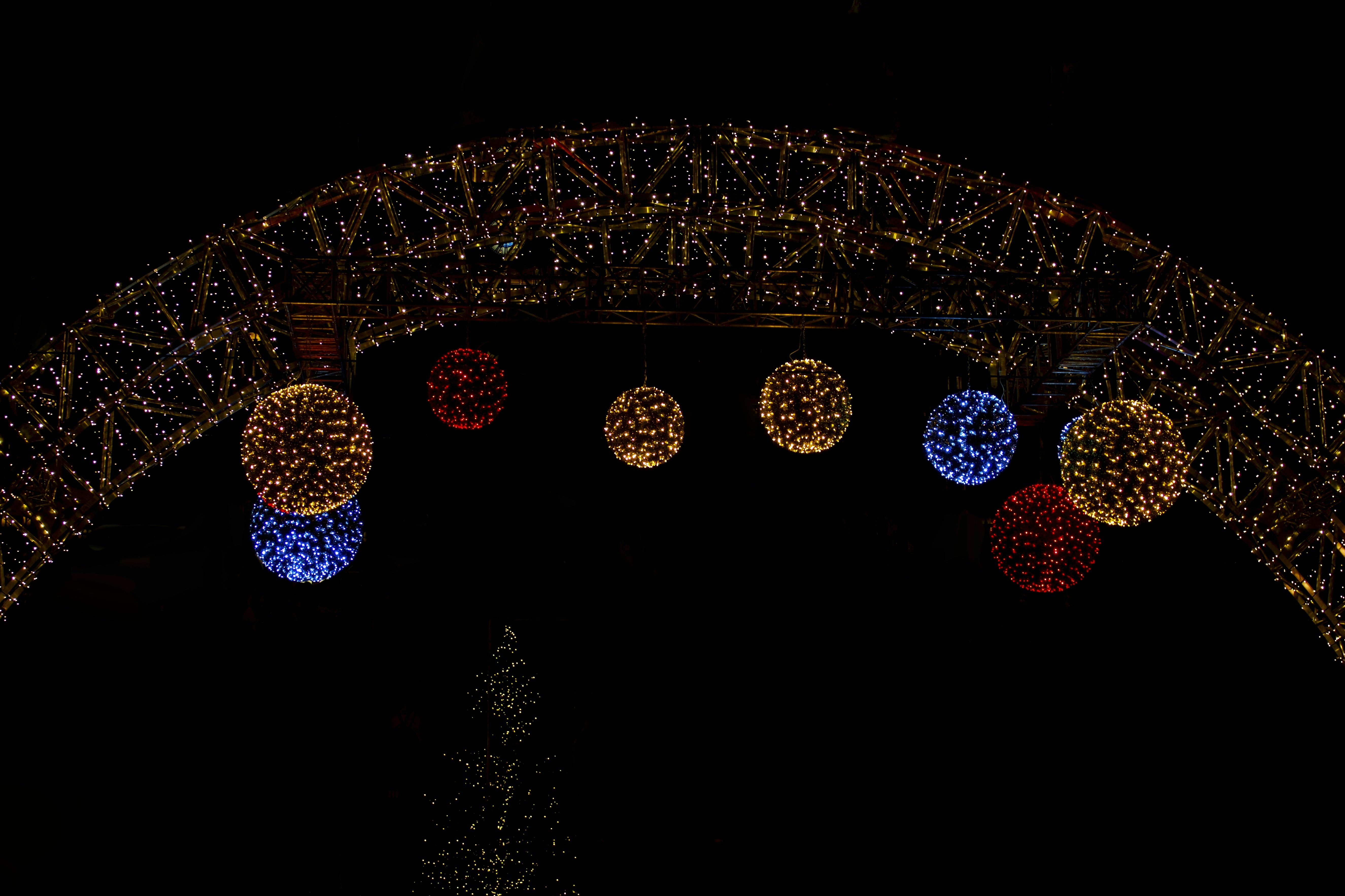 This captivating image showcases a beautifully illuminated arch adorned with vibrant, spherical Christmas lights in hues of blue, red, and gold. The dark night sky provides a perfect contrast, enhancing the glow of the lights and creating a magical, festive atmosphere. The composition highlights the symmetry and elegance of the arch, making it visually striking and enchanting.