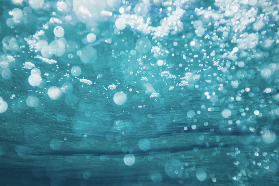 Turquoise underwater scene filled with floating bubbles and shimmering light. The water surface ripples softly near the top.