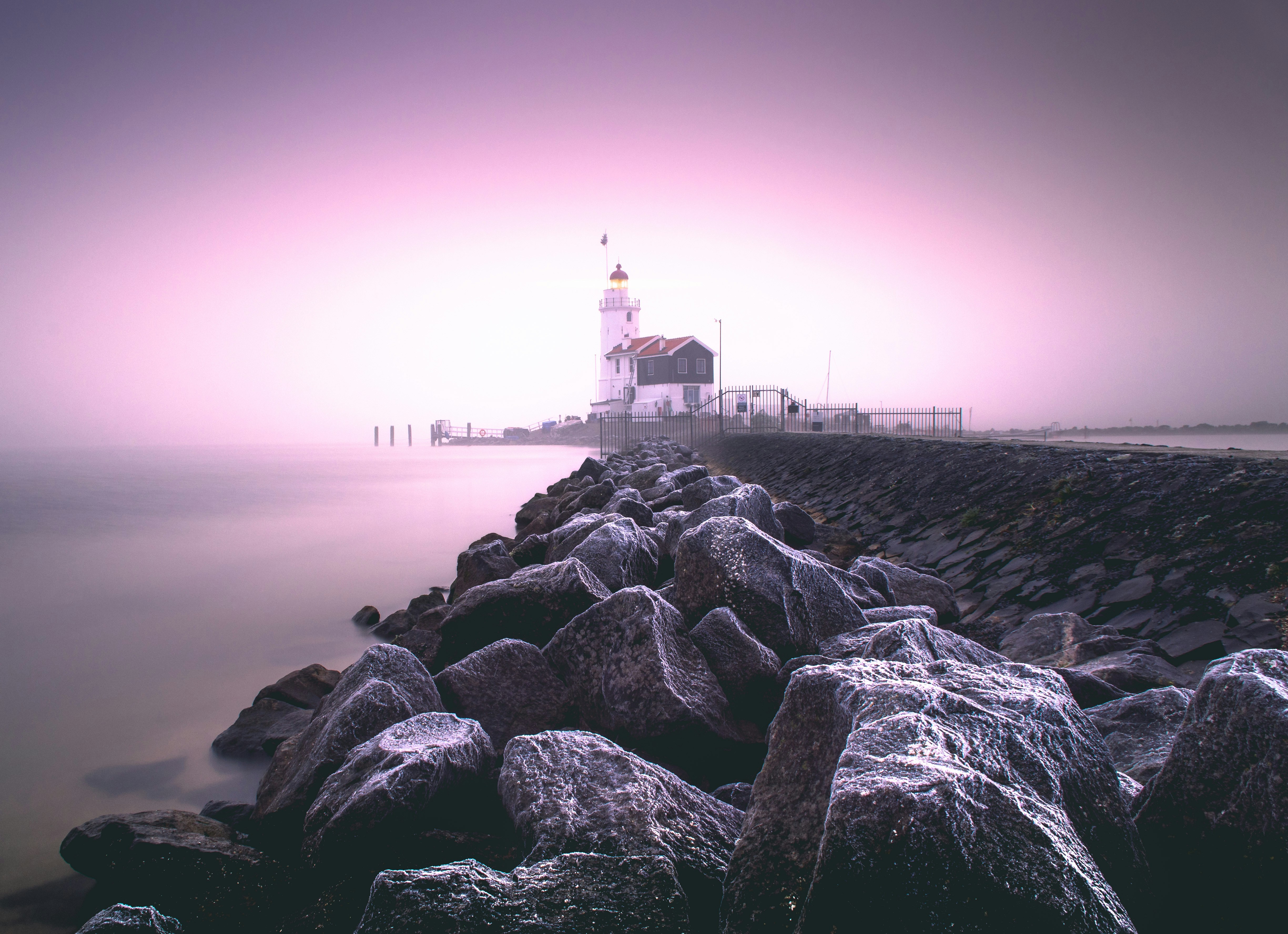 Lighthouse shrouded in mist with rocky shoreline in the foreground under a soft, purple-tinged sky.