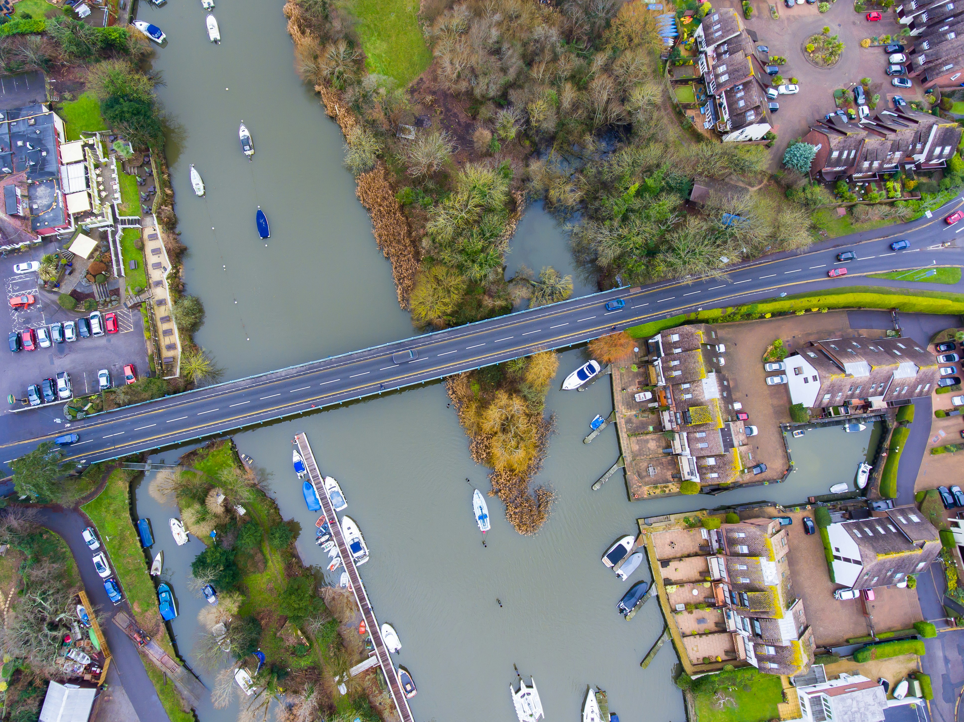 Aerial view of a bridge spanning a river with boats, surrounded by lush greenery and urban structures.