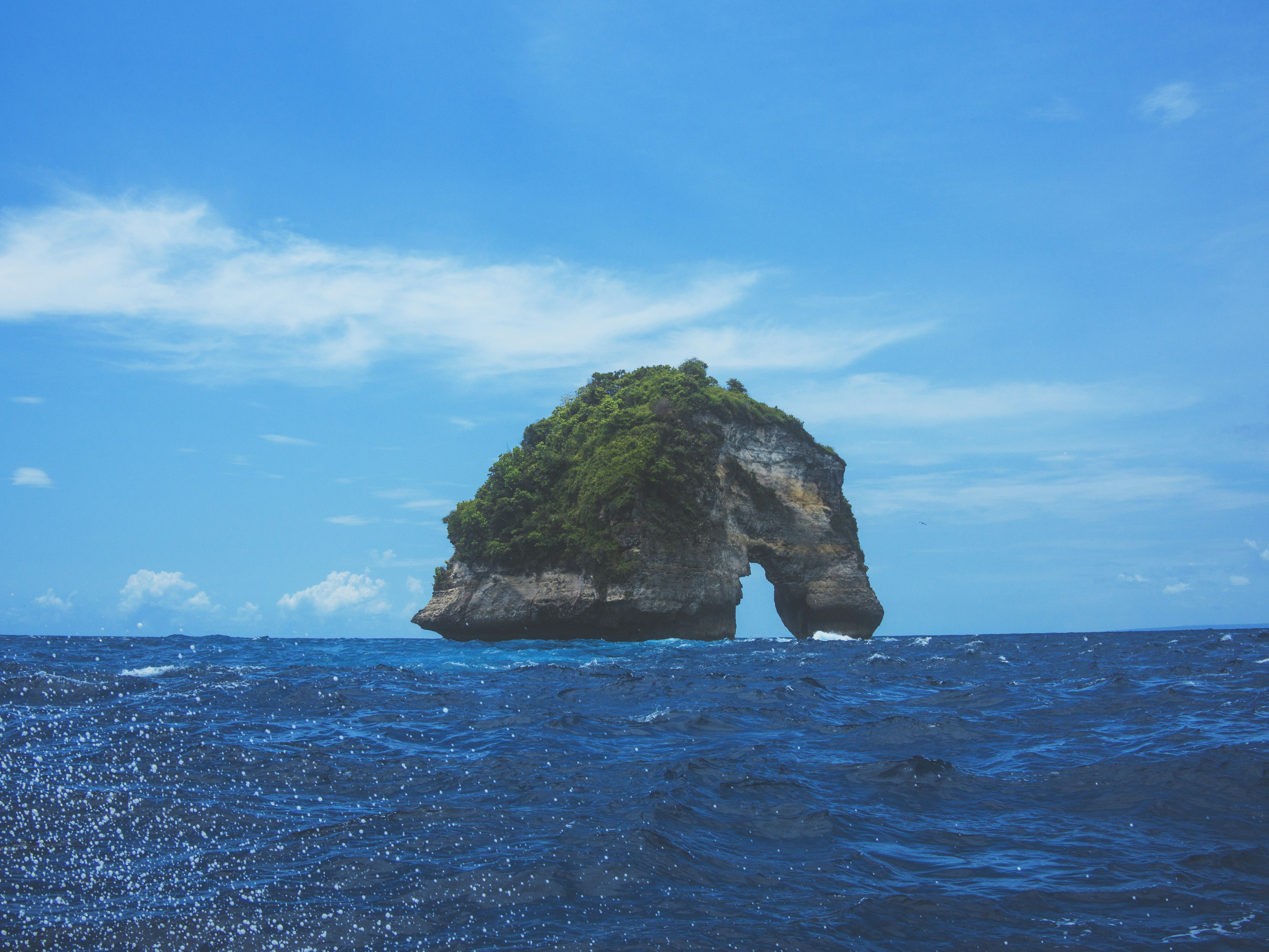 Rocky arched island surrounded by deep blue ocean beneath a sky dotted with clouds.