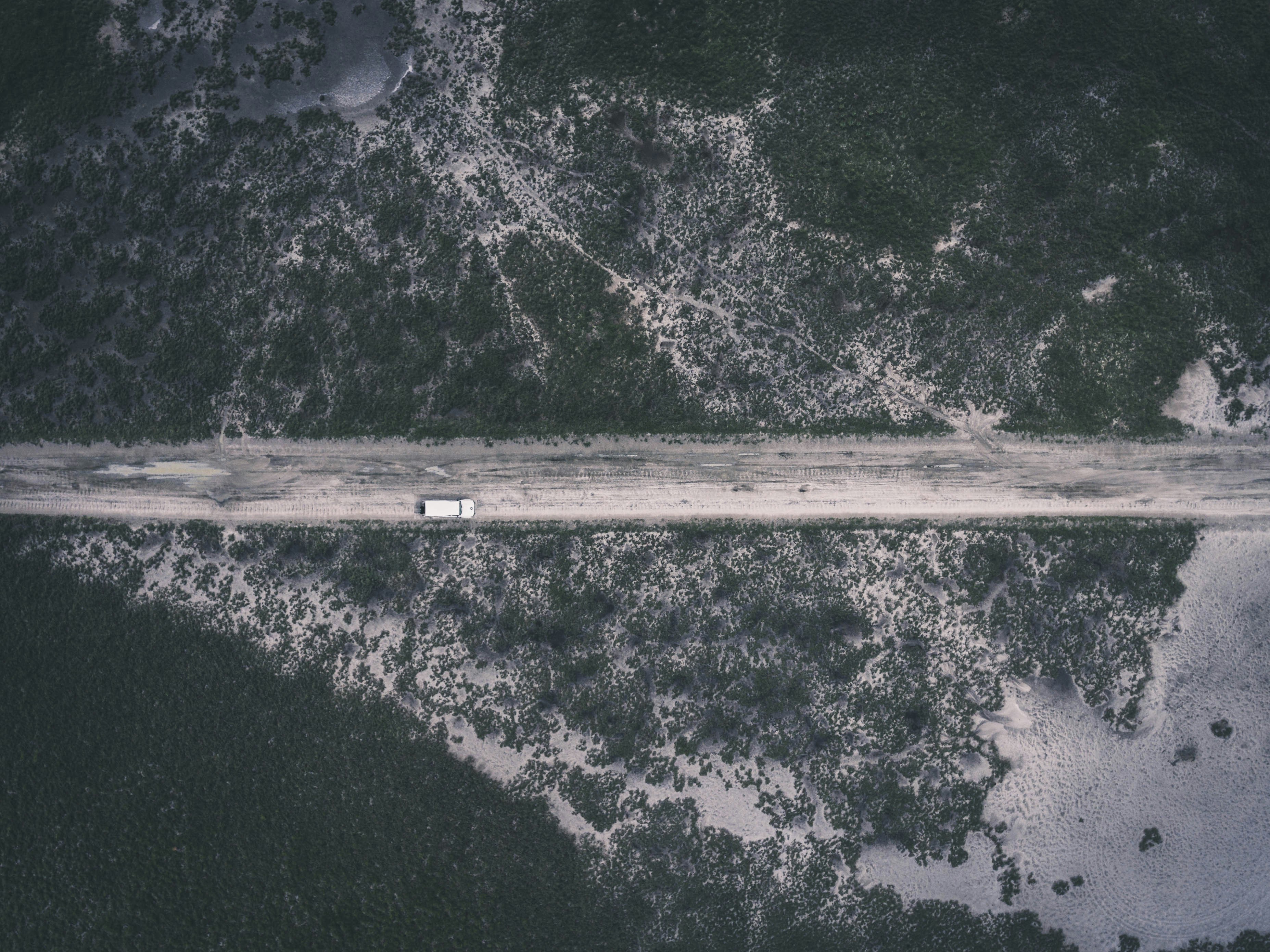 Aerial view of a lone vehicle on a dirt road surrounded by lush greenery and textured terrain. The scene captures the essence of isolation in nature.