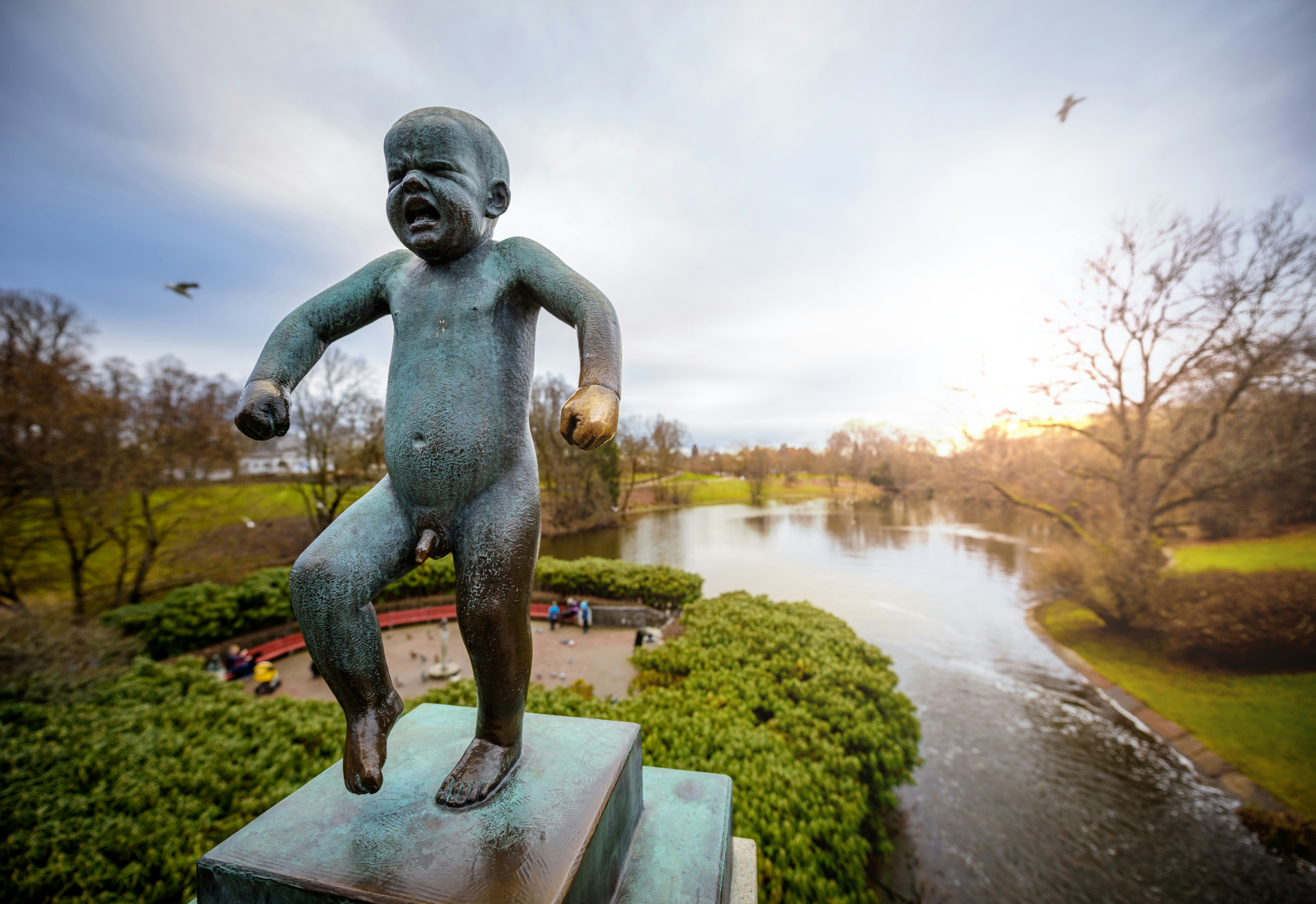 Bronze statue of a child stands prominently overlooking a serene park landscape with a river and winter trees.