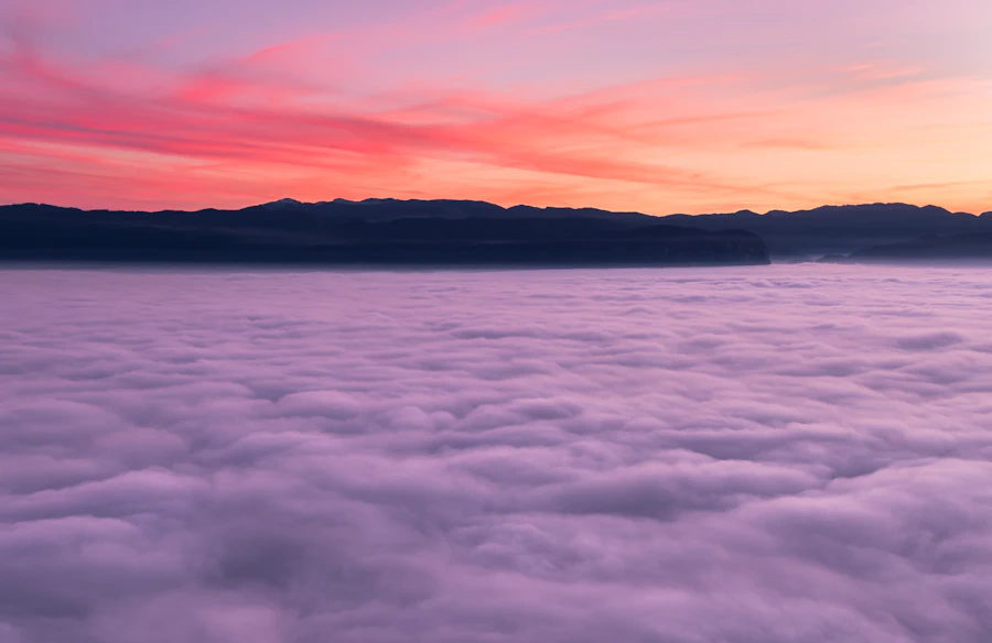 Cottony sea of clouds glows with pink-orange dawn light, a distant mountain ridge silhouettes the horizon.