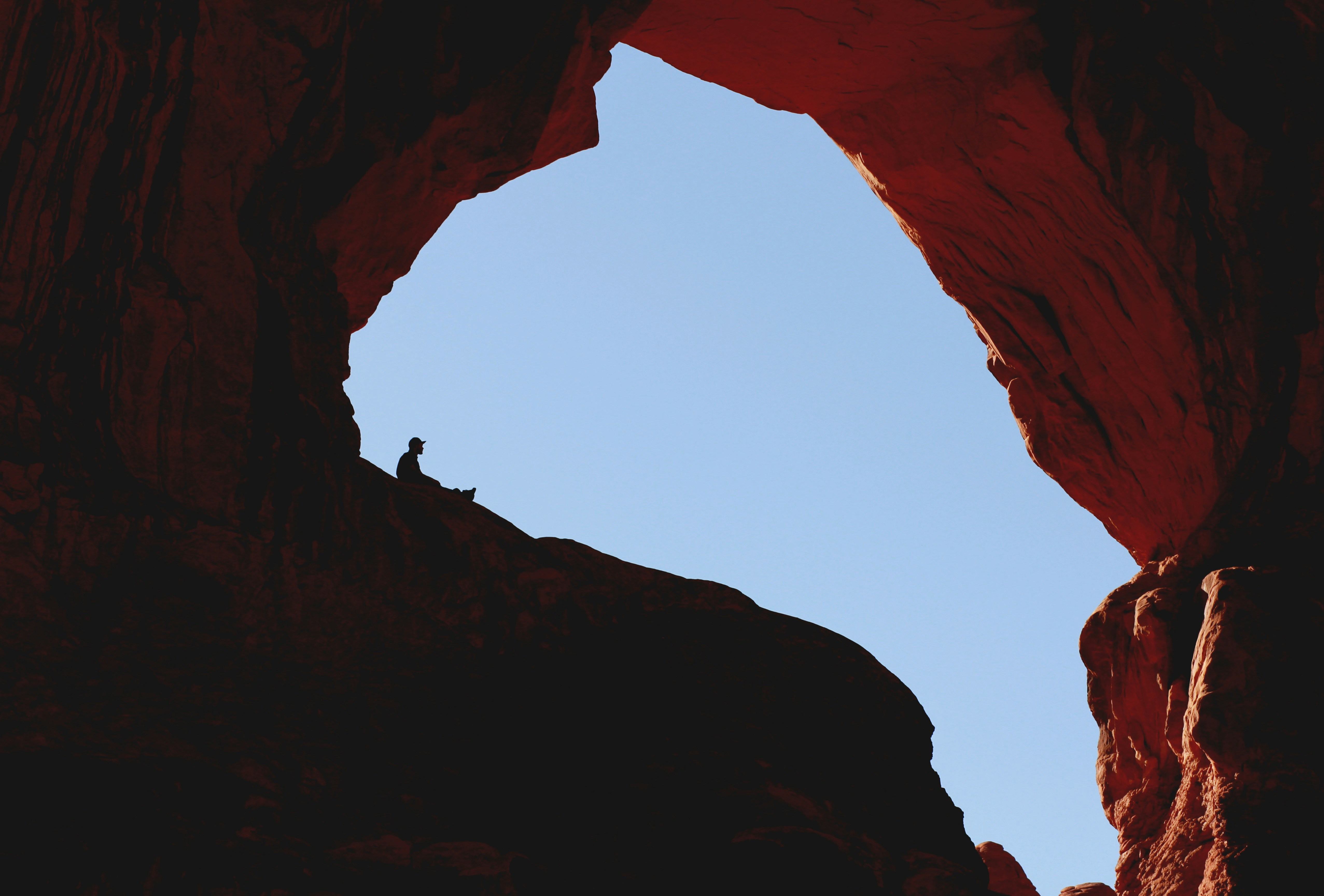 Silhouette of a person standing under a large, natural rock arch against a clear blue sky.