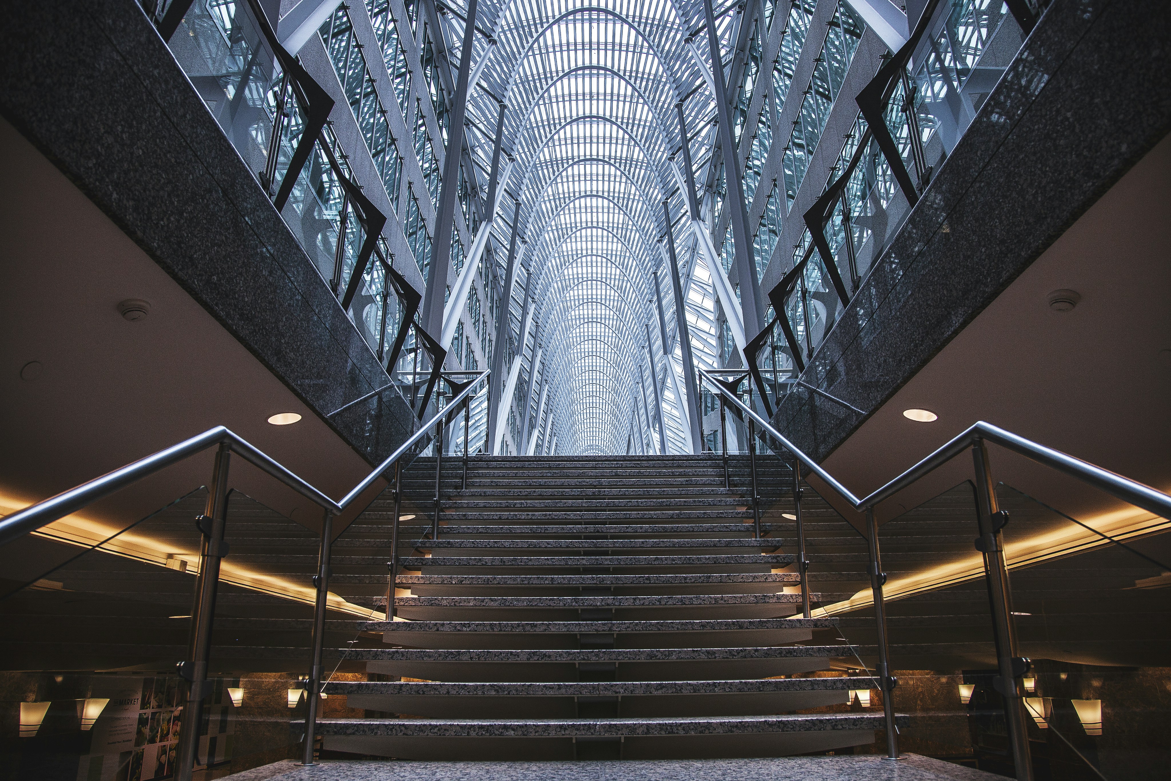 Staircase leading upwards in a modern architectural space, framed by glass and steel structures. The design emphasizes light and symmetry.