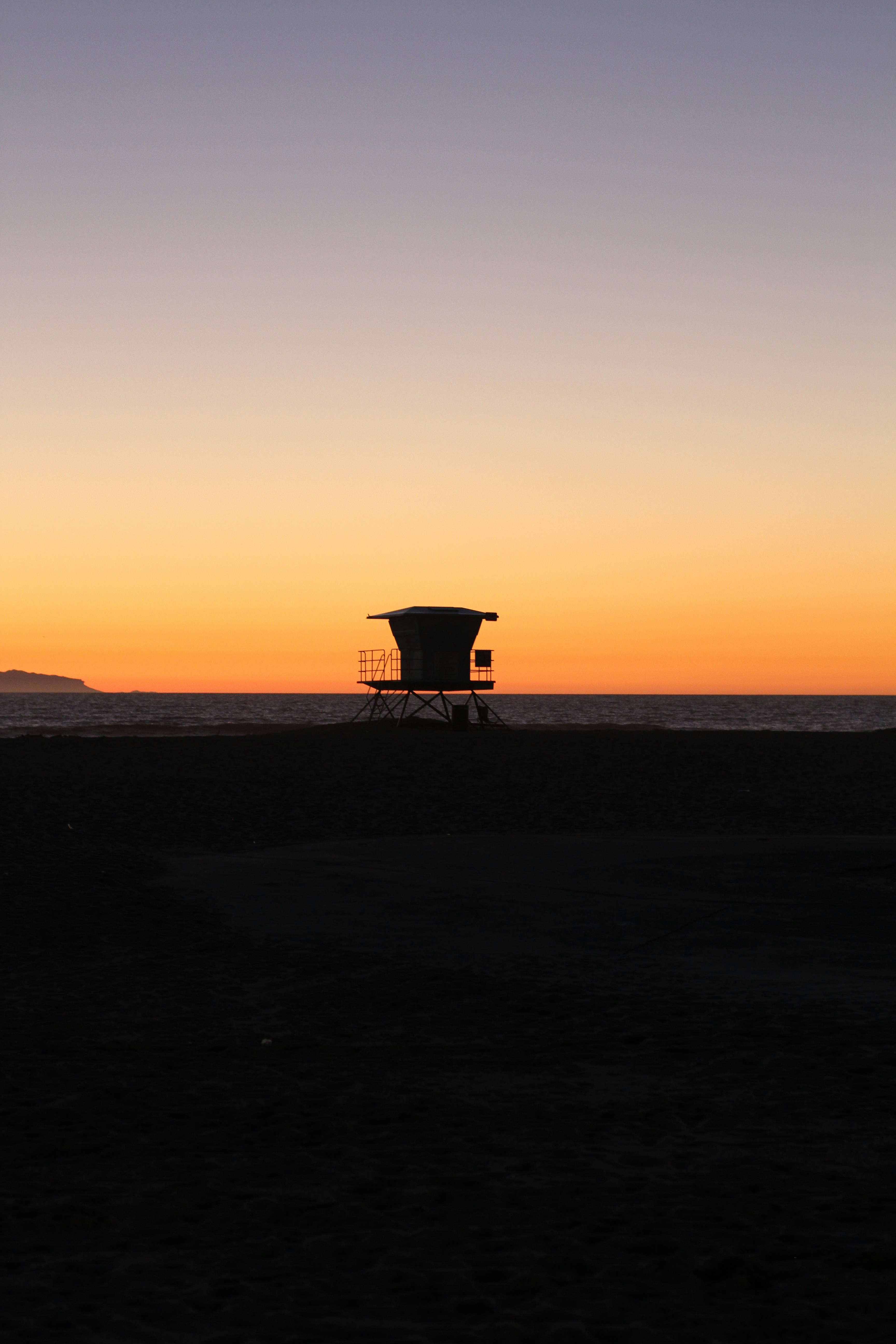 Silhouetted lifeguard tower against a gradient sunset sky, overlooking the calm ocean waves. The scene evokes a sense of tranquility and watchfulness.