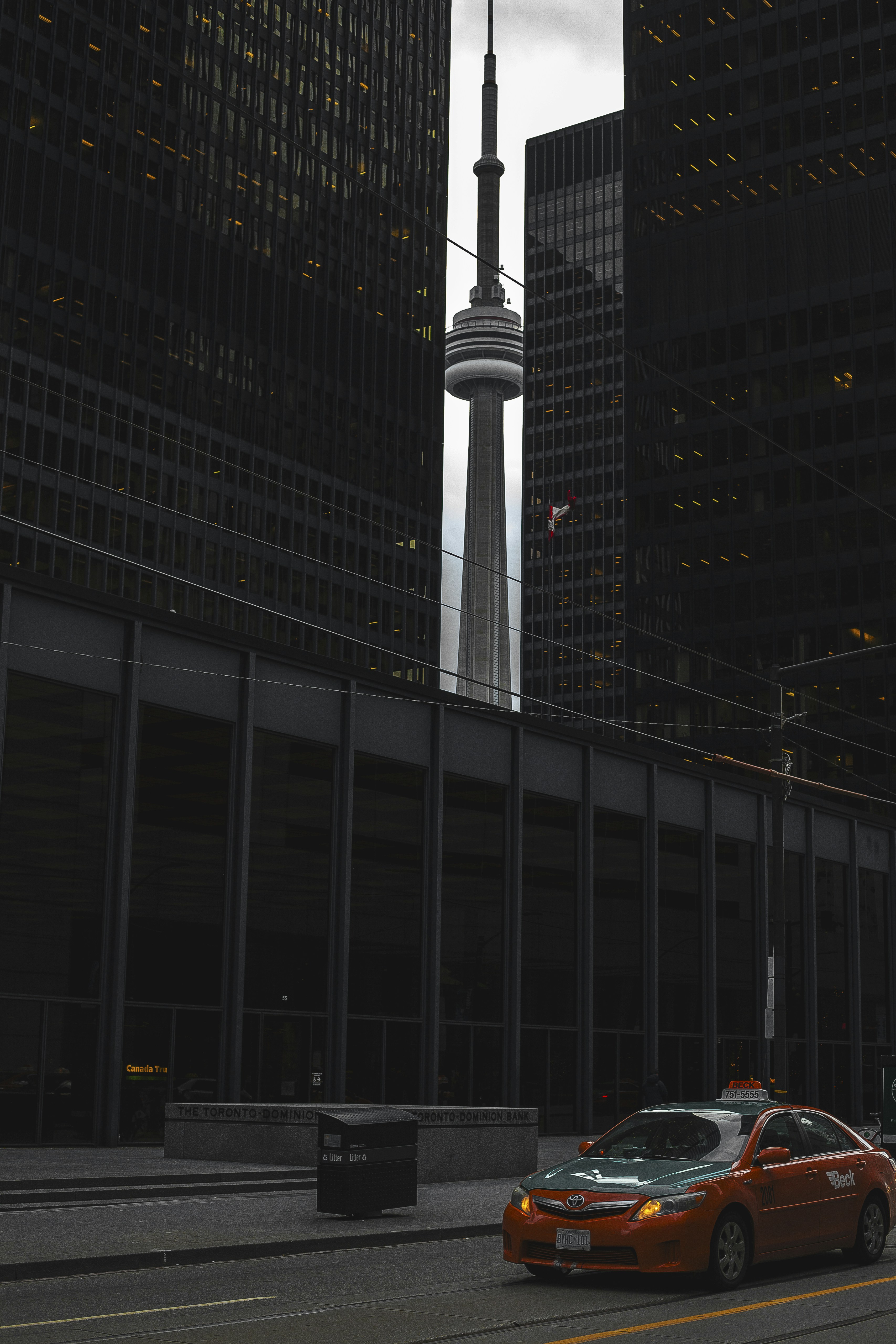 CN Tower rises dramatically between towering skyscrapers in a bustling city scene, with an orange taxi in the foreground.
