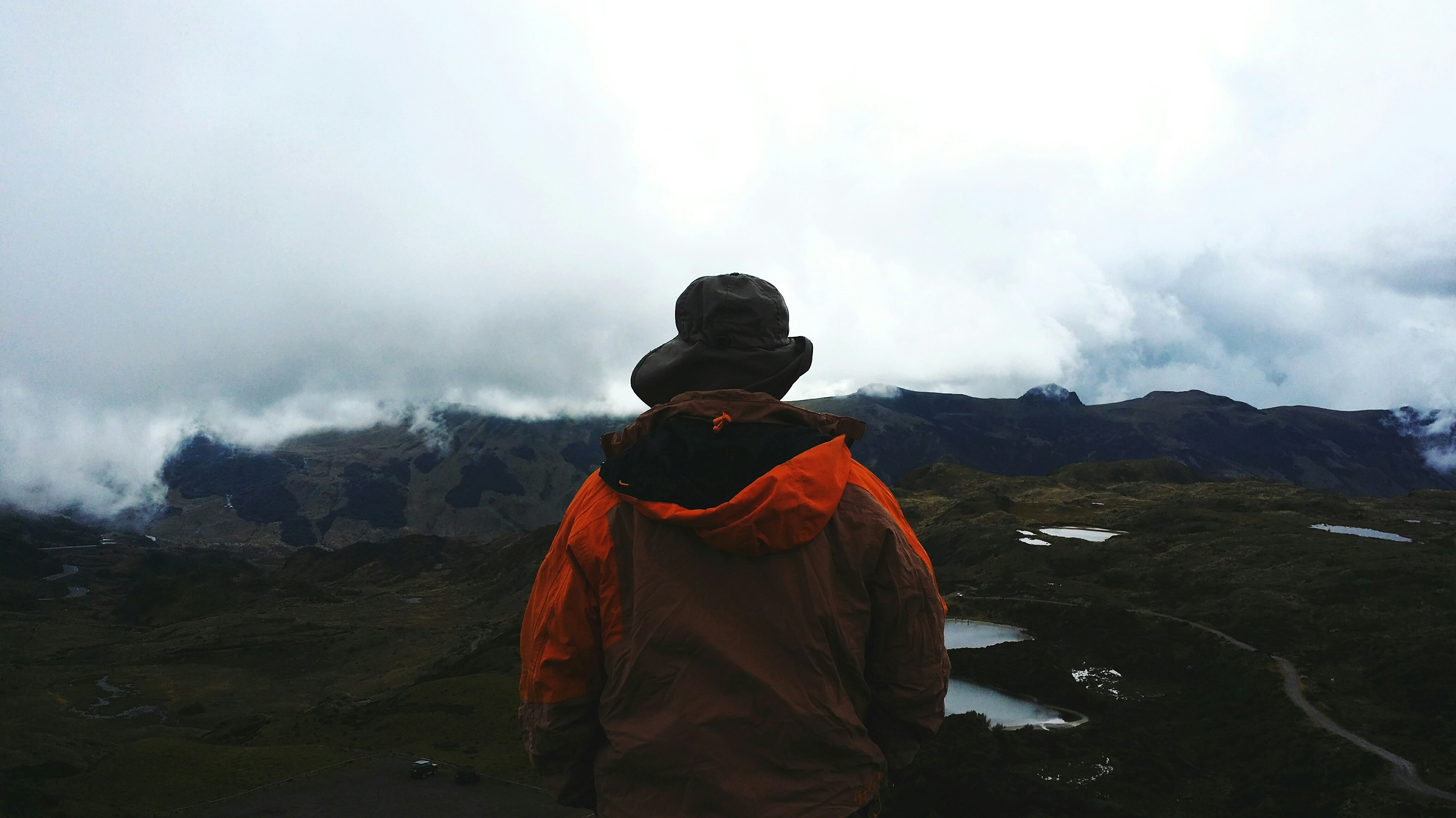 Person in an orange jacket and hat gazing at a mountainous landscape under a cloudy sky.
