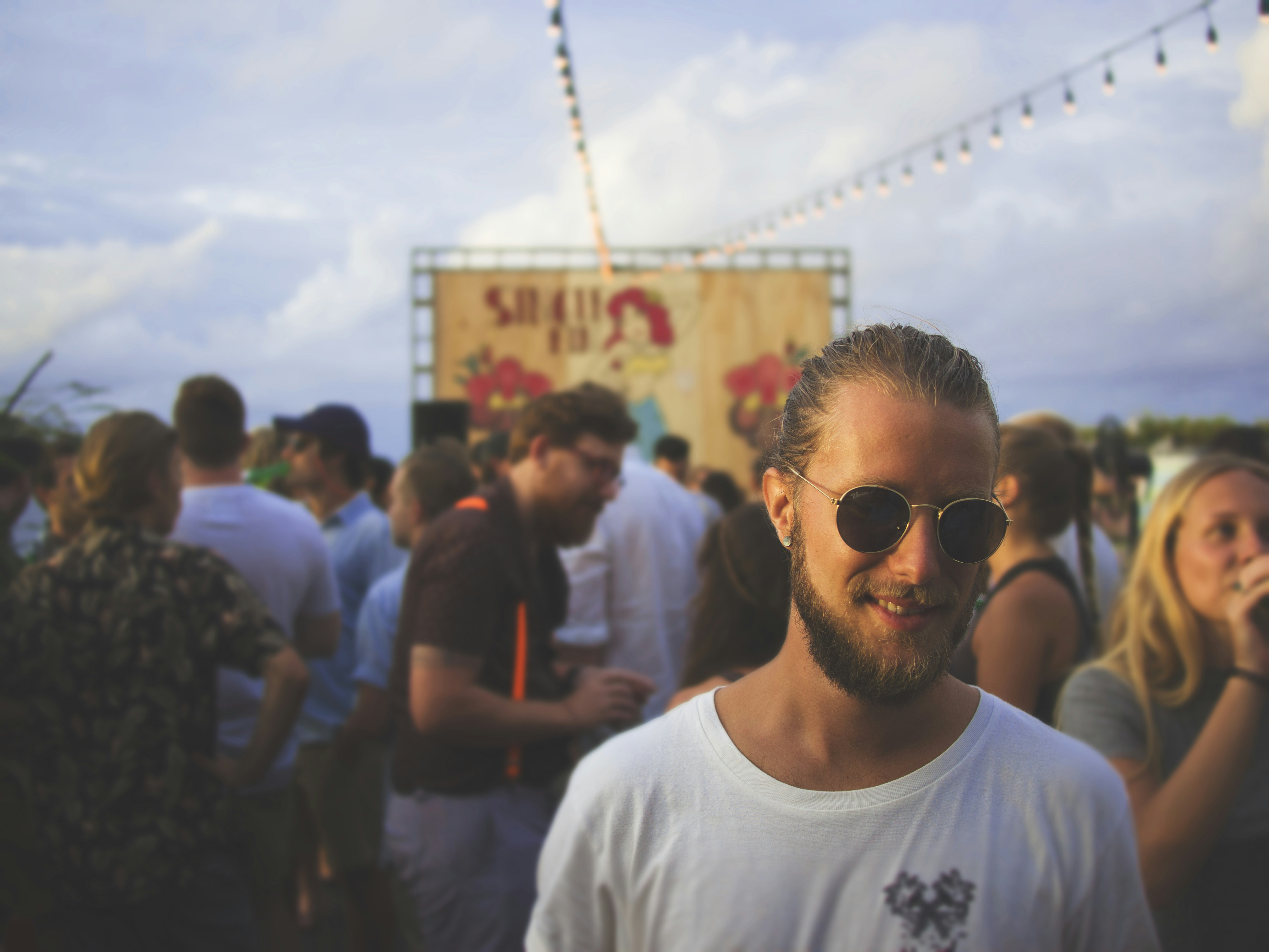 Man in sunglasses enjoying an outdoor festival with a crowd, string lights, and a colorful backdrop.