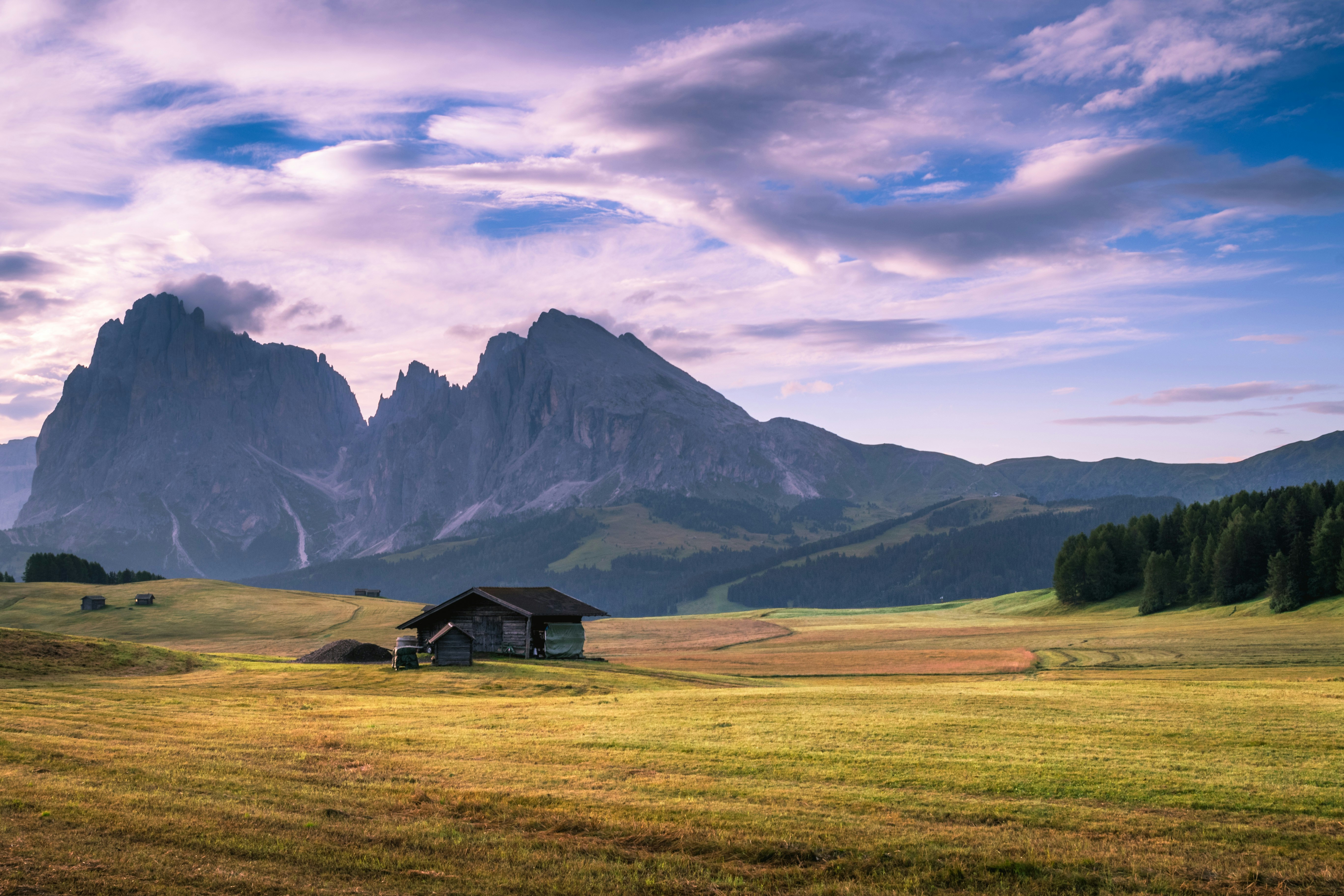 Rustic cabin nestled in a vibrant meadow with majestic mountain backdrop under a dynamic sky.
