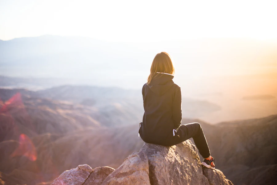 Person in a dark jacket sits on a rocky outcrop overlooking layered mountains at sunset. Warm light flares across the horizon behind them.