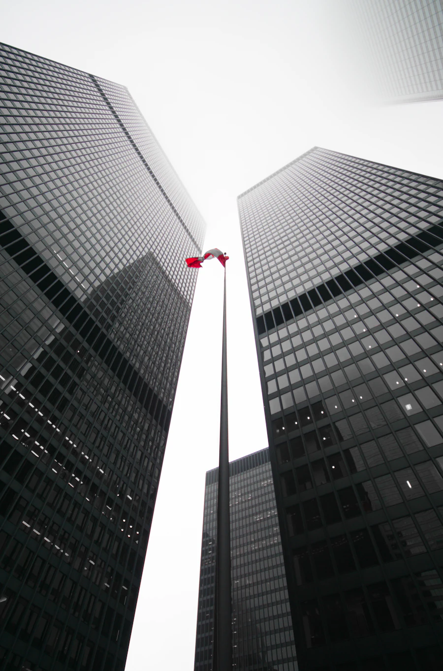 Looking up between two tall glass office towers with a red-and-white flag on a central pole against a bright overcast sky.