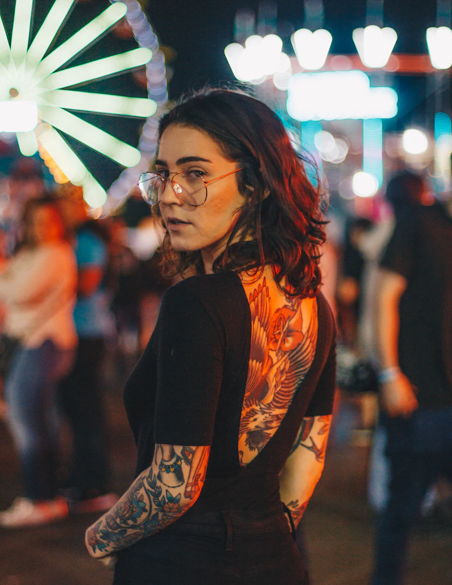 Tattooed woman in glasses looking back over her shoulder at a nighttime fair, with colorful neon lights and blurred crowds behind her.