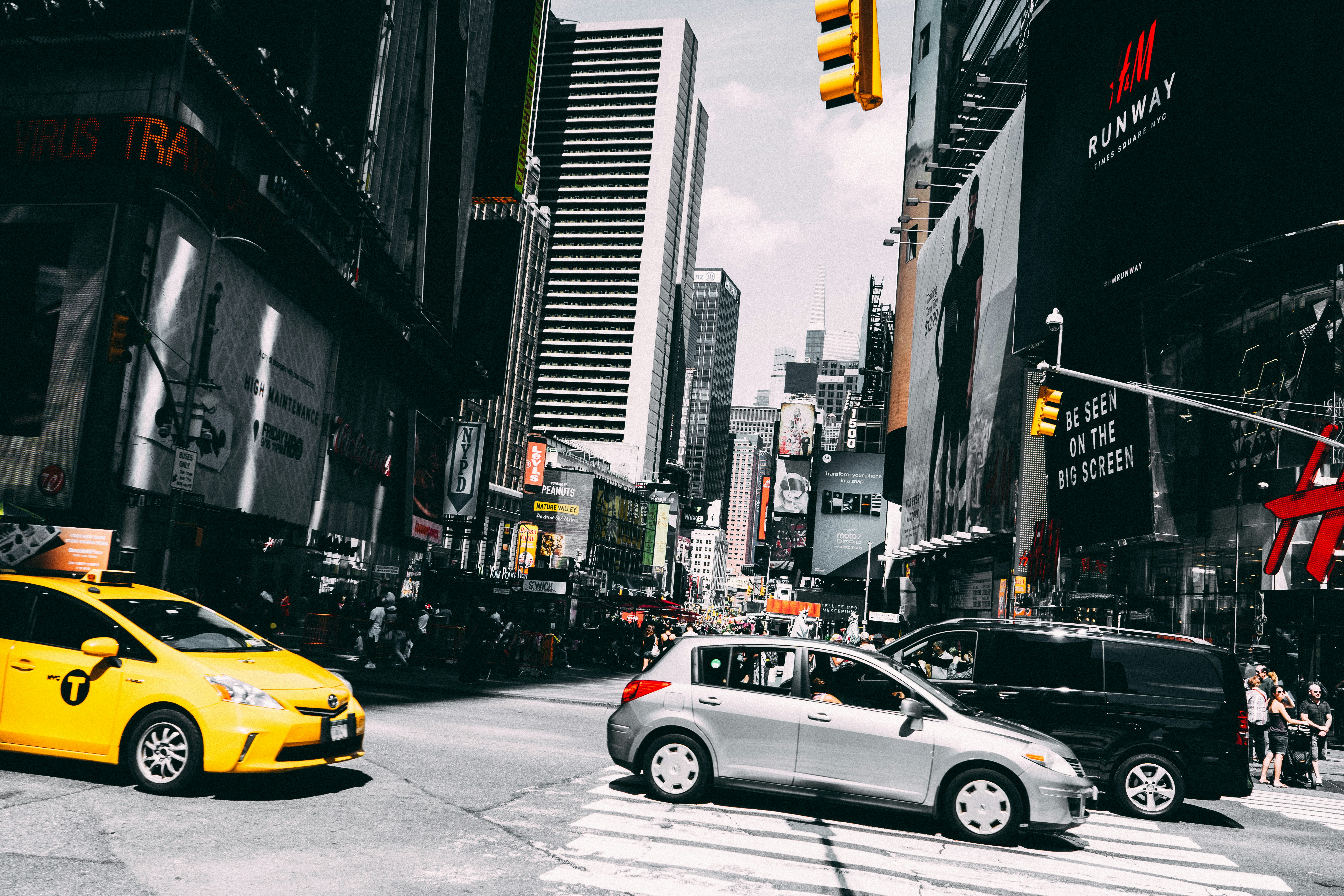 Yellow taxi and silver car crossing a busy New York City intersection under towering skyscrapers.