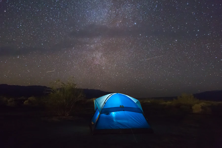 A glowing blue tent sits in a dark desert landscape beneath a star-filled night sky and the Milky Way. Low shrubs and distant hills silhouette the horizon.