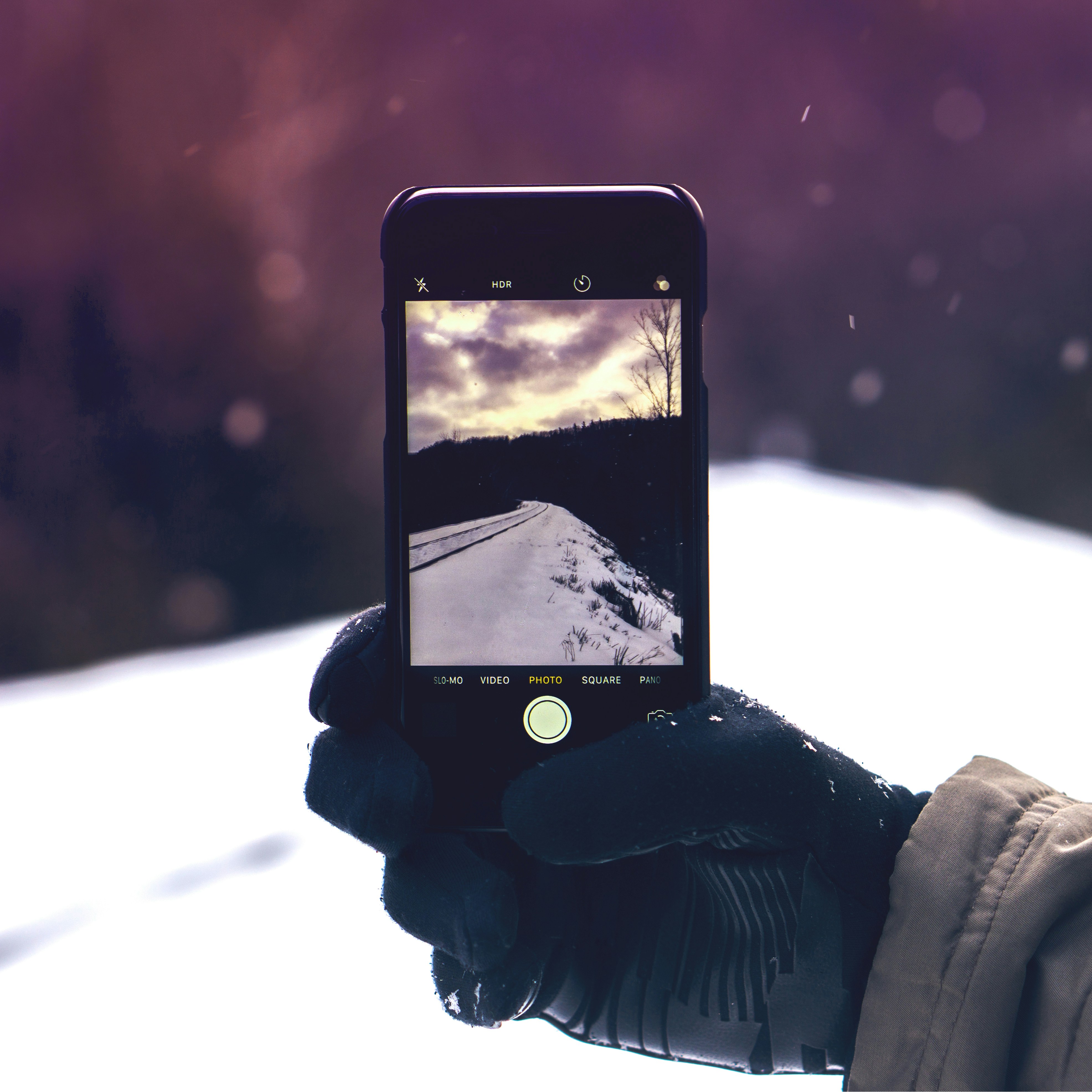A gloved hand holds a smartphone capturing a snowy landscape with a winding path and dramatic clouds. The scene highlights the intersection of technology and nature.