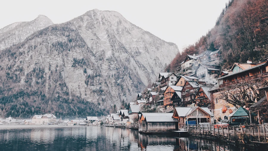 A small cluster of lakeside houses clings to a steep mountainside beneath snow-dusted peaks, with calm water reflecting the village and cliffs. Bare winter trees frame the right edge.