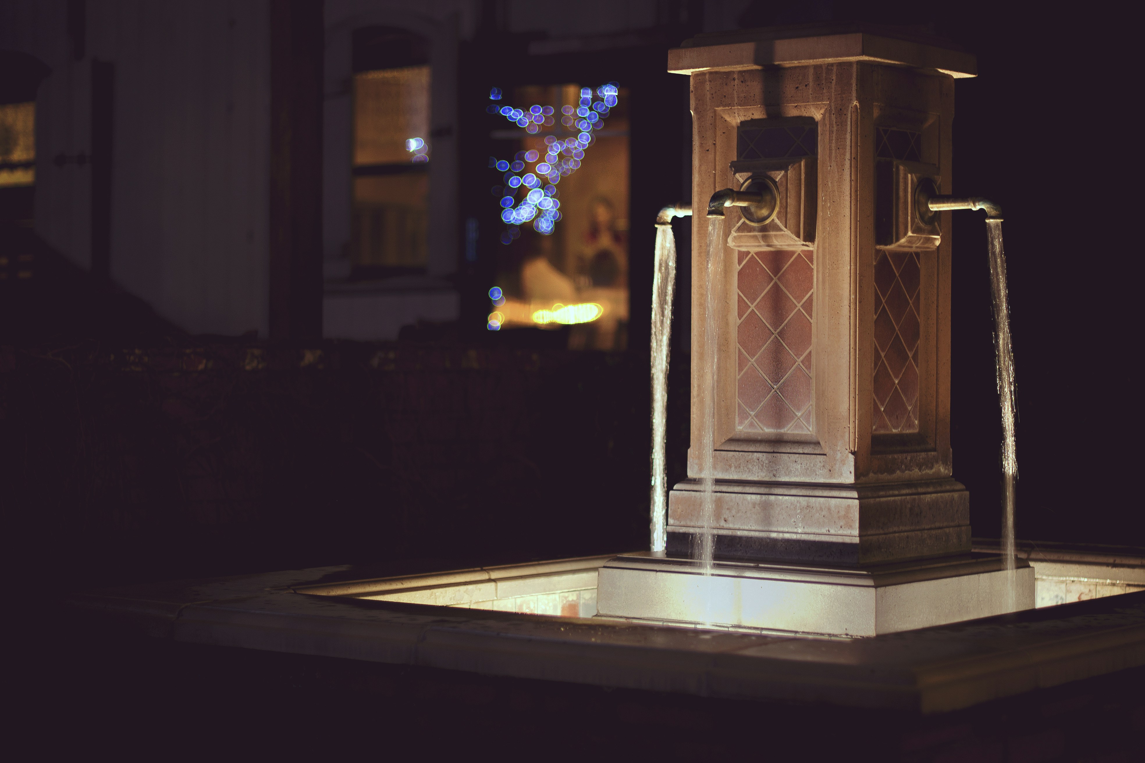 Elegant fountain with flowing water illuminated by soft light, set against a blurred backdrop of festive lights.