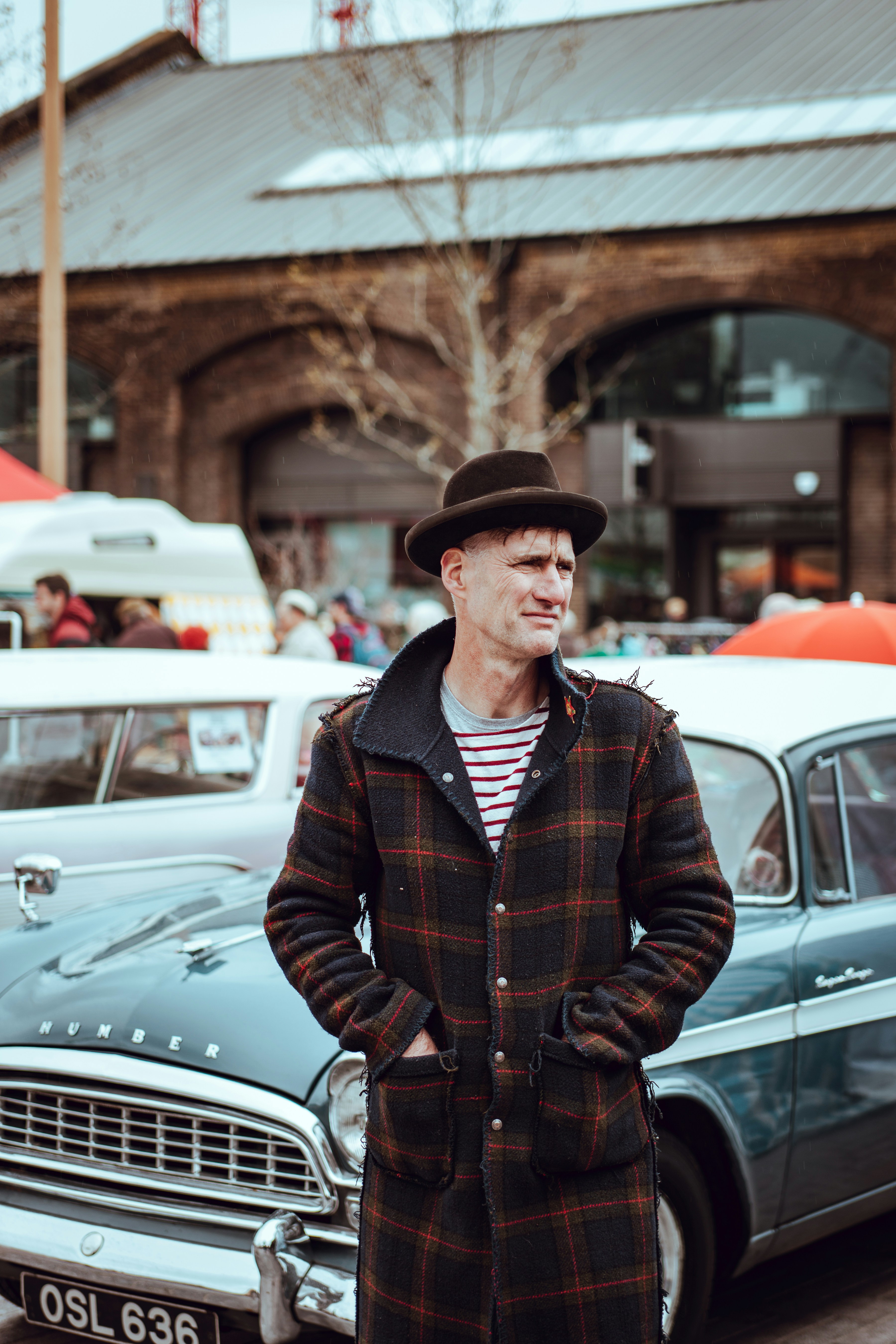 A man in a plaid coat and hat stands in front of a classic car, surrounded by a lively market scene. The atmosphere reflects a nostalgic celebration of vintage automobiles.