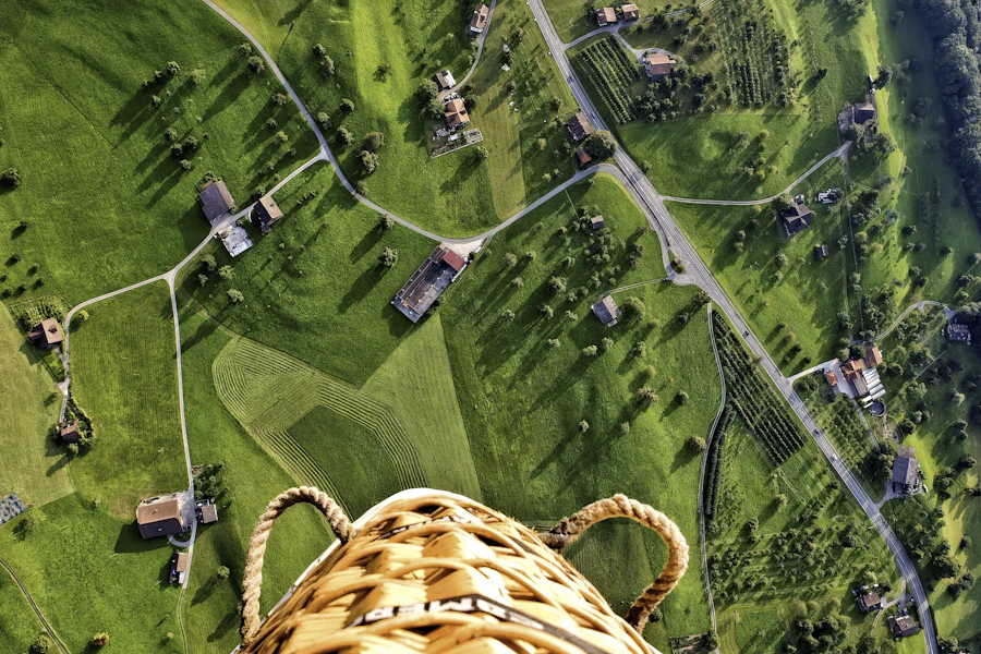 View from a hot air balloon looking down over vivid green farmland, roads, and scattered houses. The balloon basket and ropes frame the bottom edge of the scene.