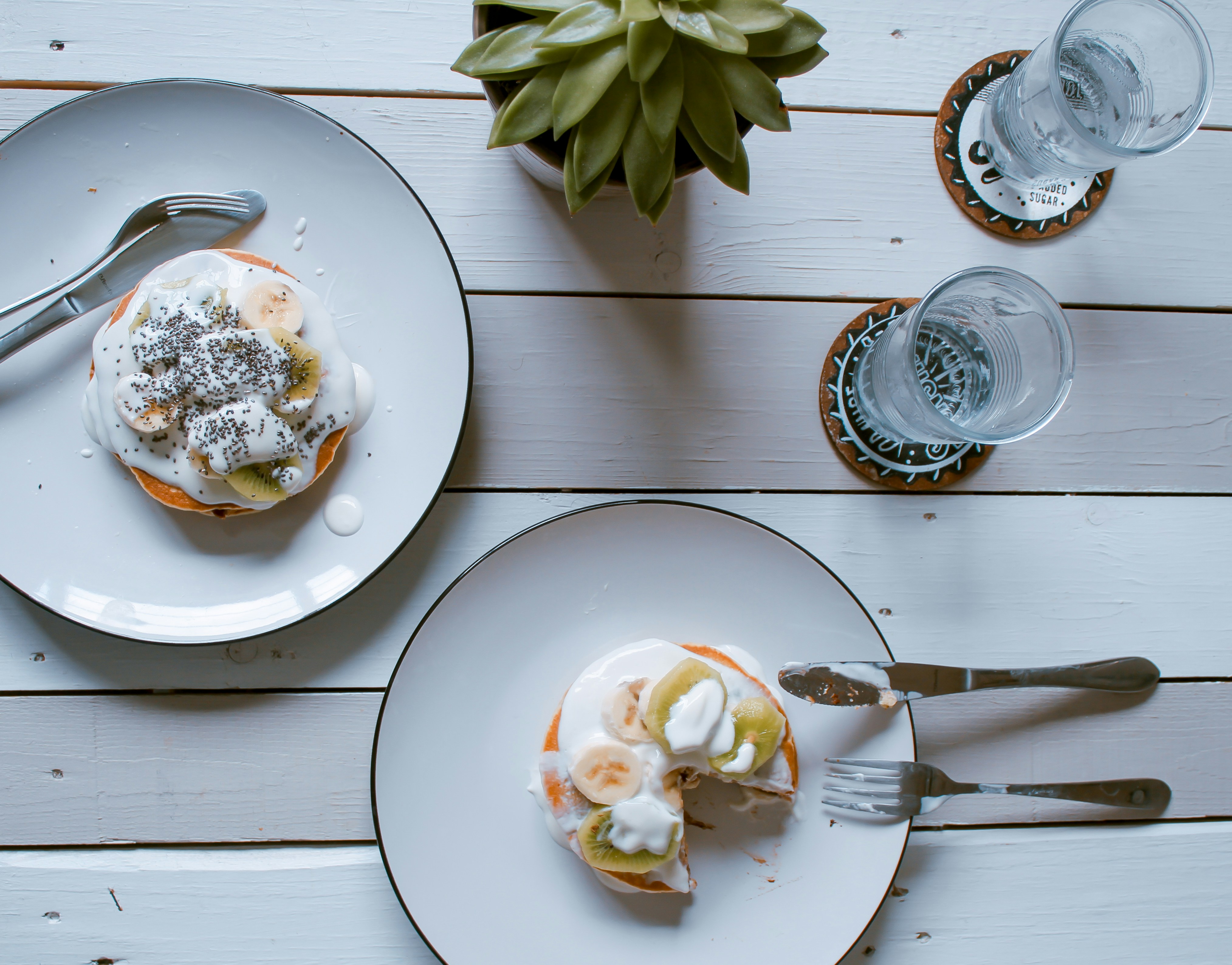 Plates of waffles topped with fruit and cream on a white wooden table alongside drinks and a small plant.