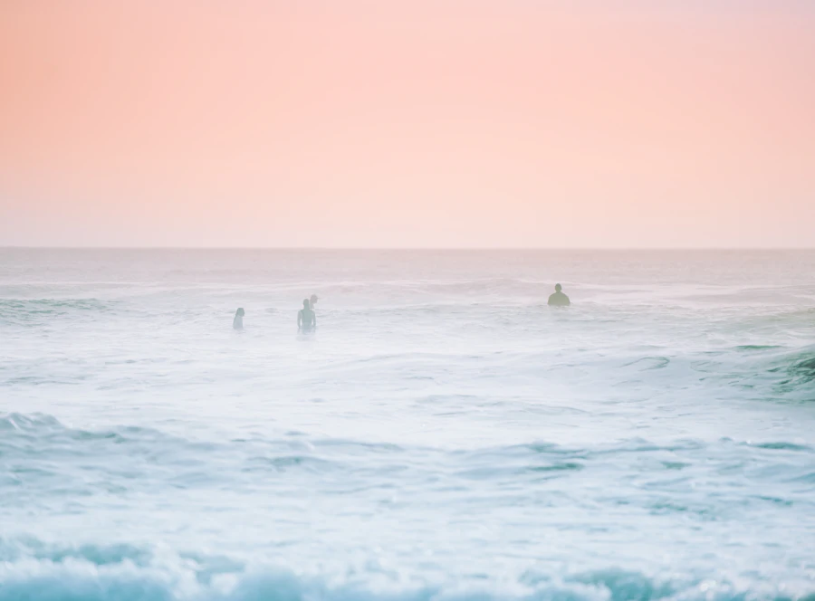 Surfers navigate gentle waves under a soft, pastel sunrise sky.