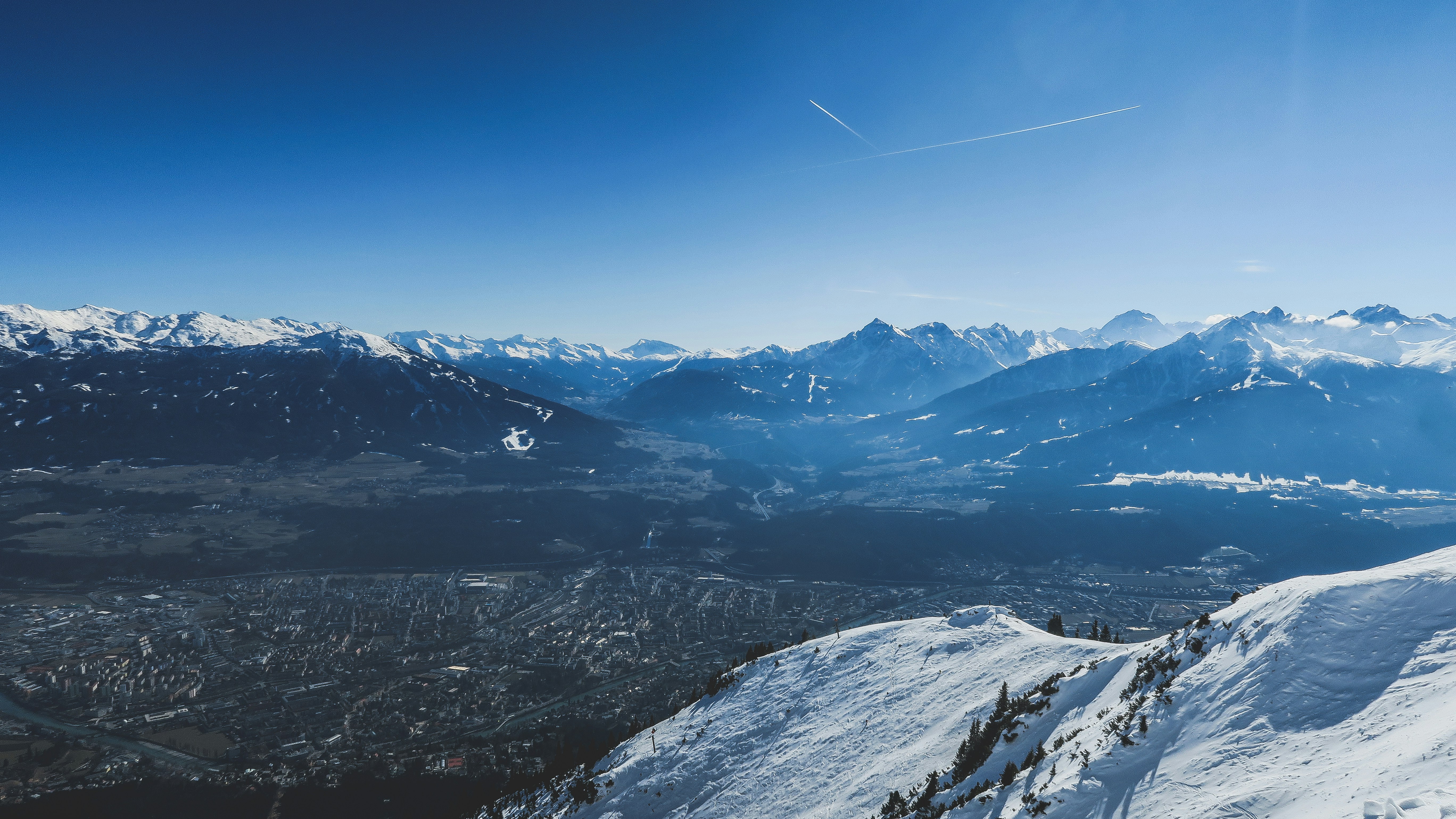 Snow-covered mountain range with a panoramic view of a valley and town below, under a clear blue sky. The scene captures the serene beauty of winter landscapes.