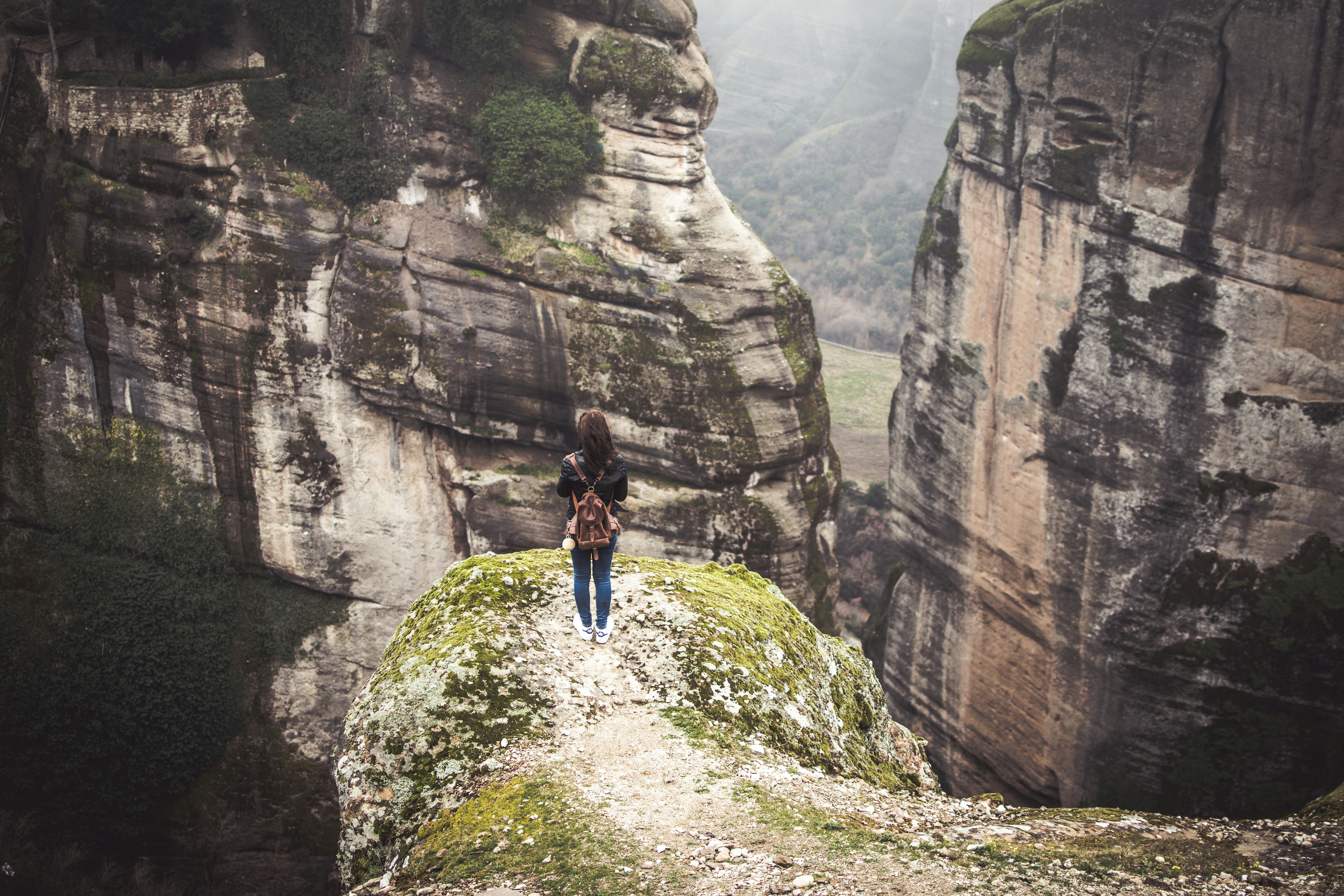 Person standing on a moss-covered rock overlooking steep cliffs at Meteora, Greece.
