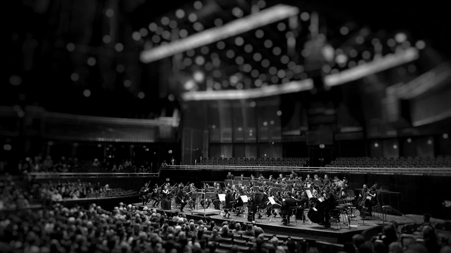 A full orchestra performs on a large stage in a modern concert hall, with the audience filling the foreground and blurred ceiling lights overhead. The scene is rendered in high-contrast black and white.