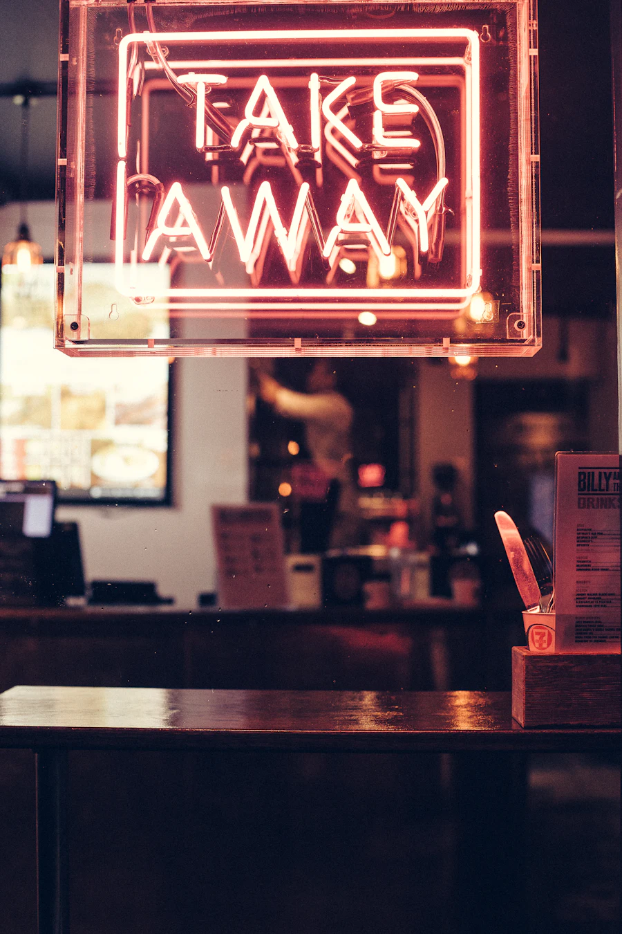 Neon 'Take Away' sign illuminating a dimly lit restaurant interior.