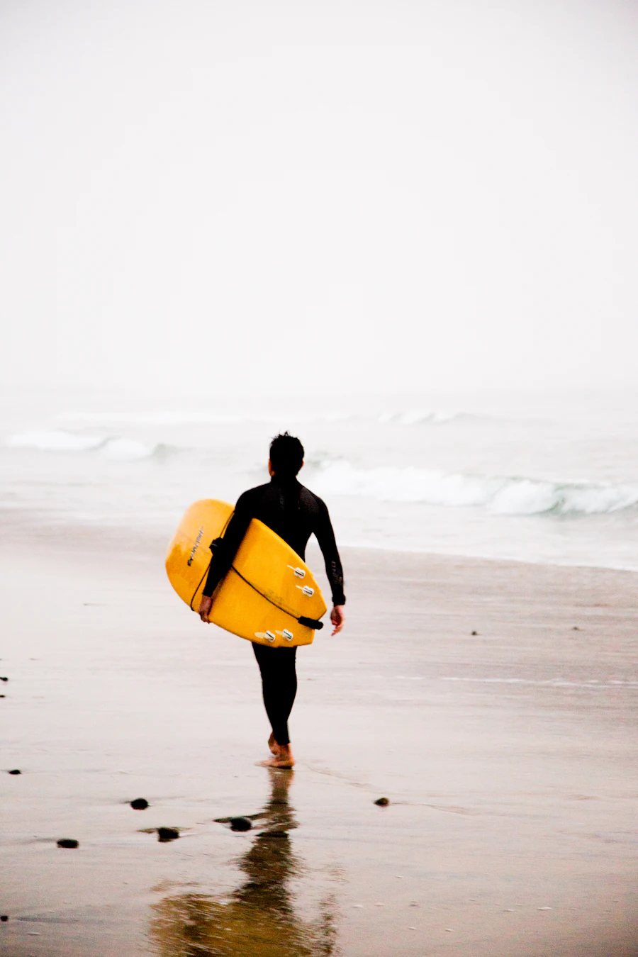A lone surfer strides along the shoreline carrying a bright yellow board, waves curling in the distance under a pale sky.