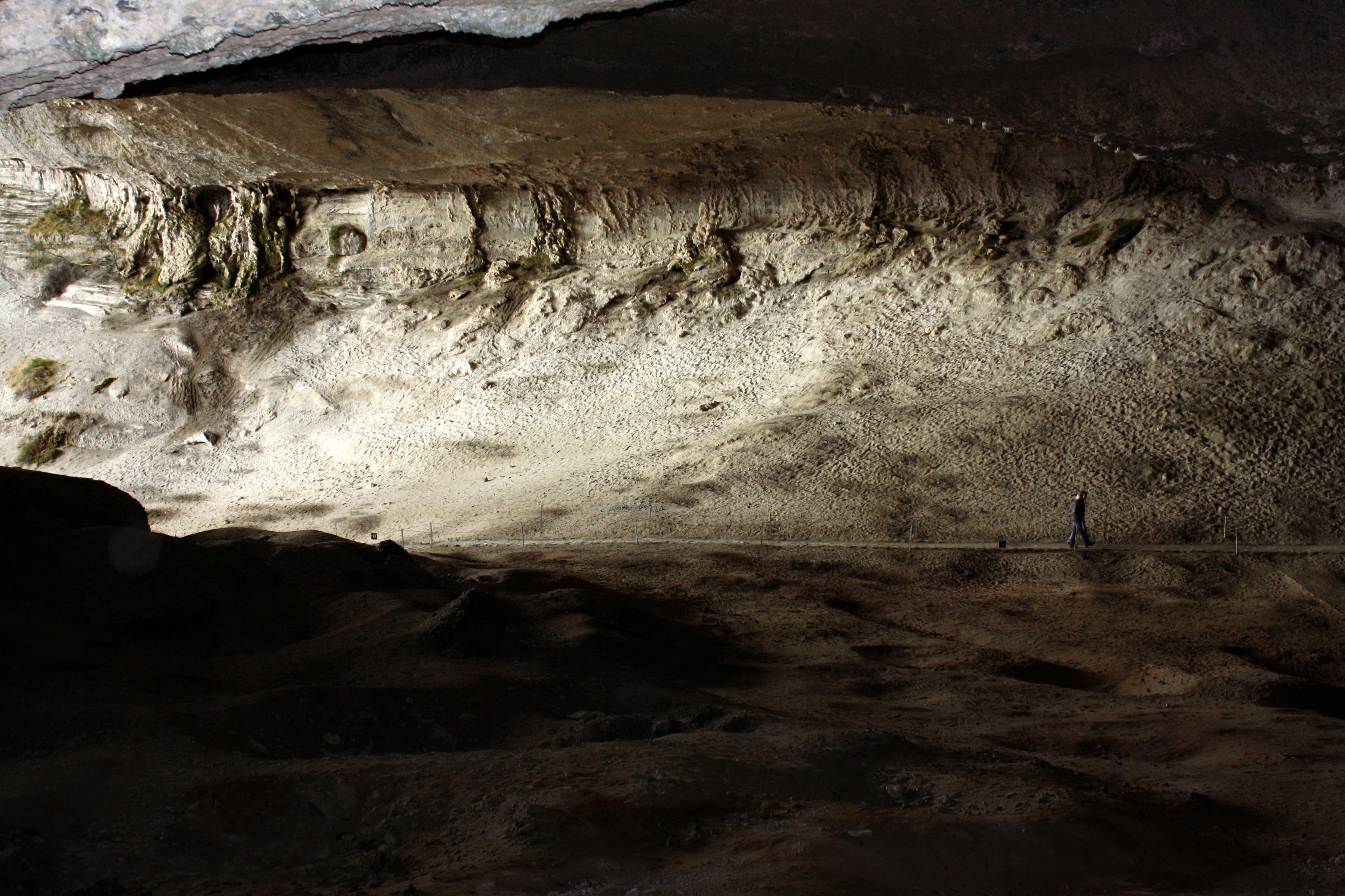 Figure walking along a narrow path inside a vast, illuminated cave with textured walls.