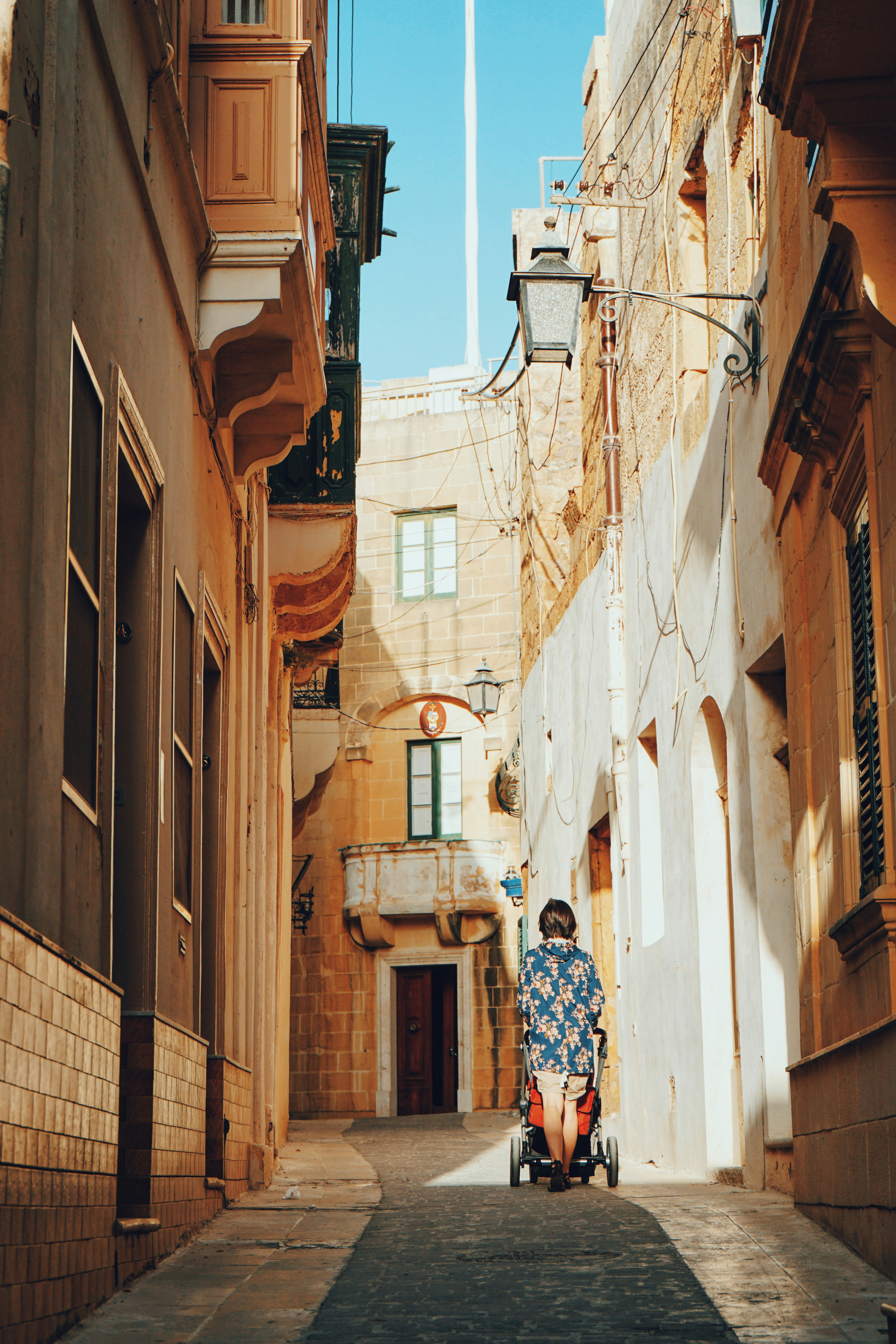 Person pushing a stroller down a narrow, sunlit alley with warm-colored buildings in Victoria, Malta.