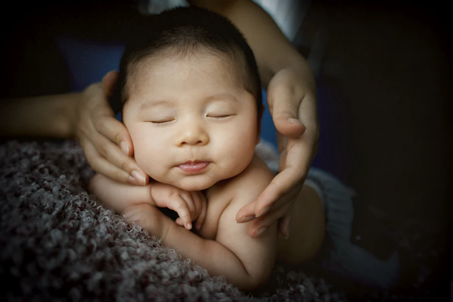 Sleeping newborn resting on a textured blanket, framed by protective adult hands. Soft, warm light and a dark background create an intimate portrait.