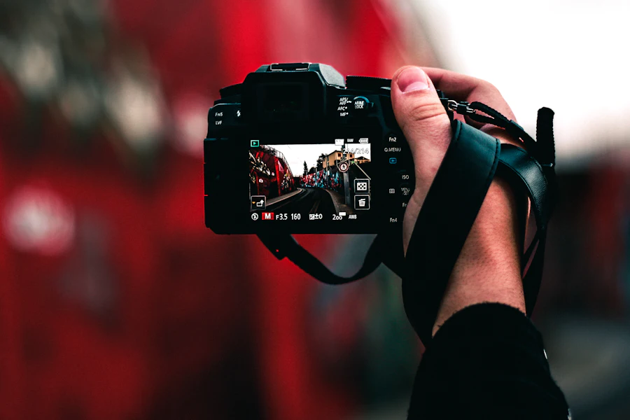 A hand holds a Canon DSLR showing a live view of a street scene, with a heavily blurred red background. The camera strap drapes across the frame.