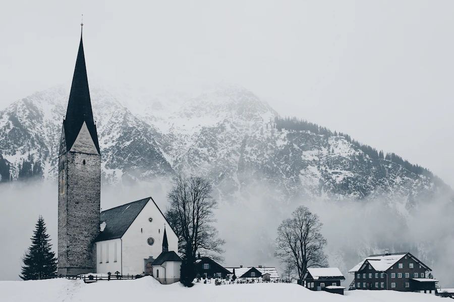 A tall church steeple rises from a snow-covered village, with bare trees and houses scattered below. Fog softens the rugged, snowy mountains in the background.