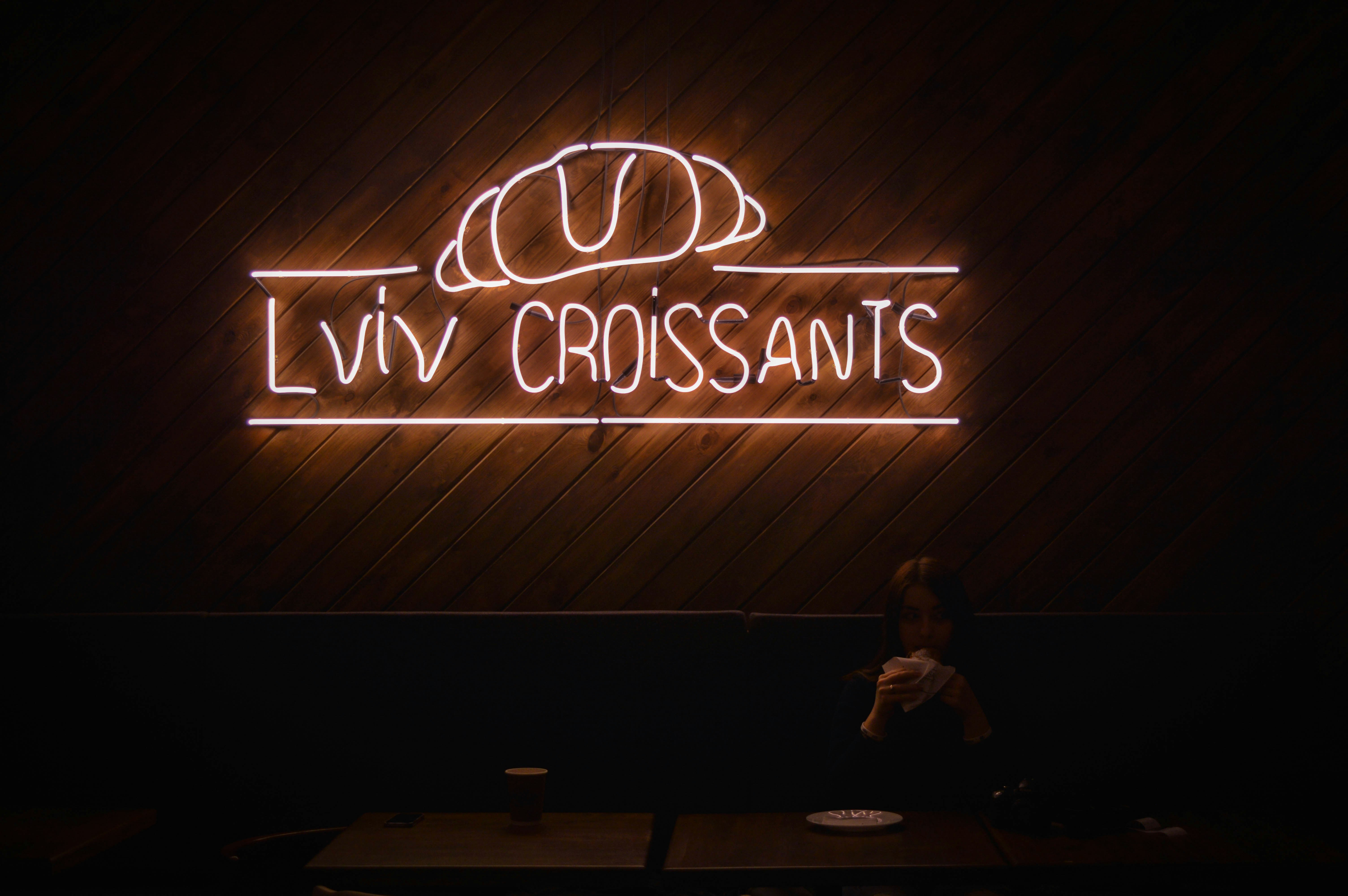 Neon sign reading 'Lviv Croissants' illuminates a dark, wood-paneled wall with a person beneath enjoying a pastry.
