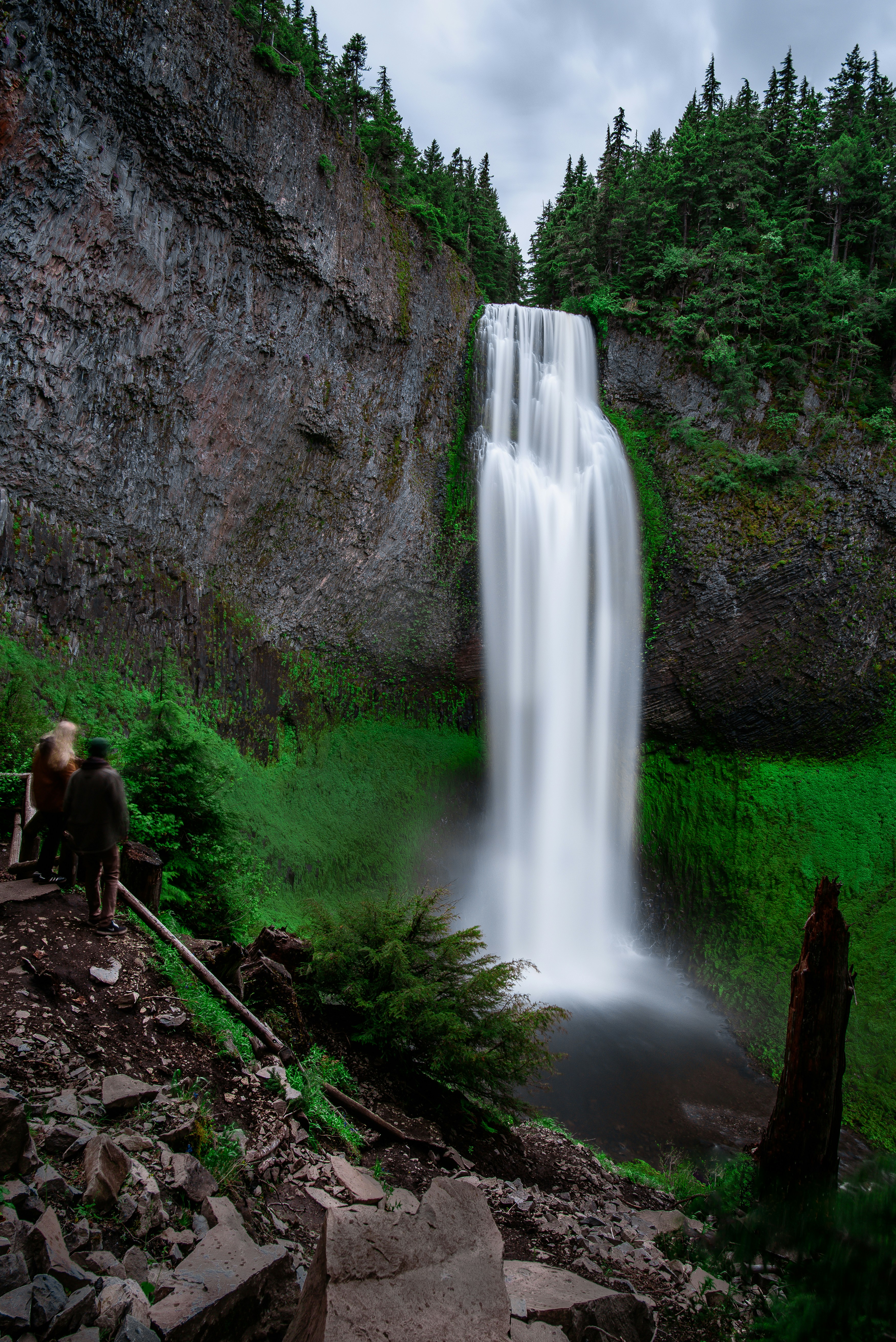 A majestic waterfall cascades down a rocky cliff, surrounded by lush greenery and moss-covered stones. Two figures stand nearby, admiring the serene beauty.