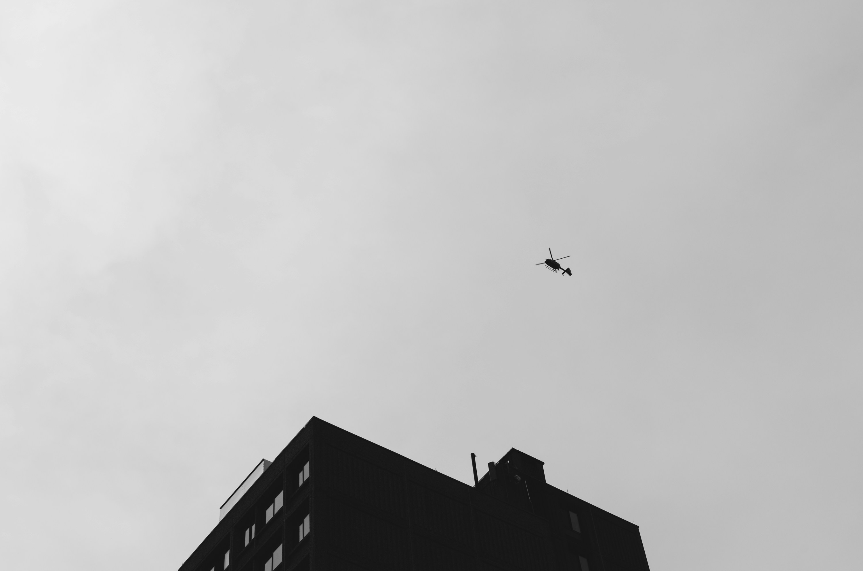 A helicopter hovers above a tall building, set against a moody, overcast sky. The stark contrast of the dark structure emphasizes the aircraft's presence.