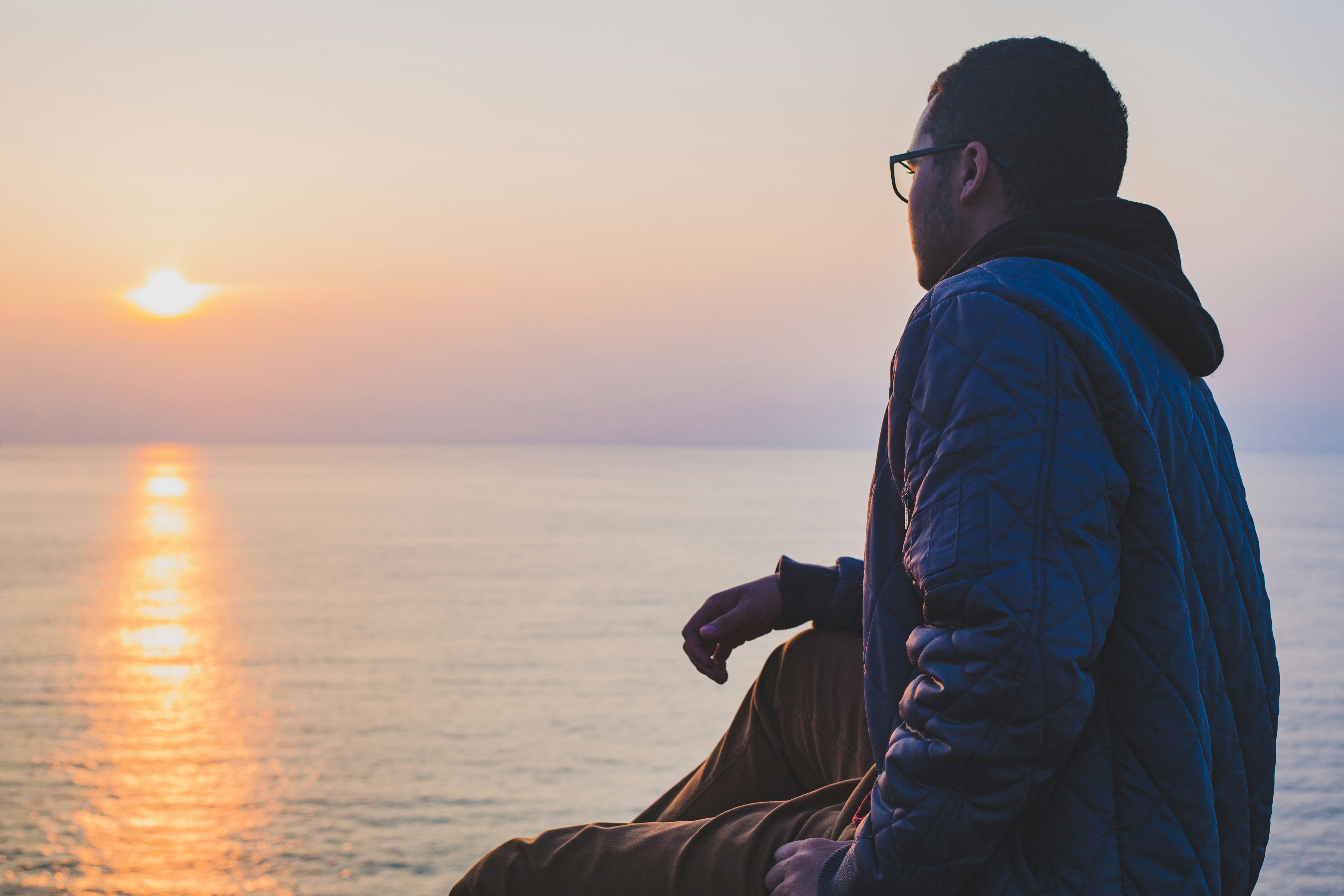 A contemplative figure gazes at the horizon as the sun sets over the tranquil sea, creating a serene atmosphere. The scene captures the essence of quiet reflection.